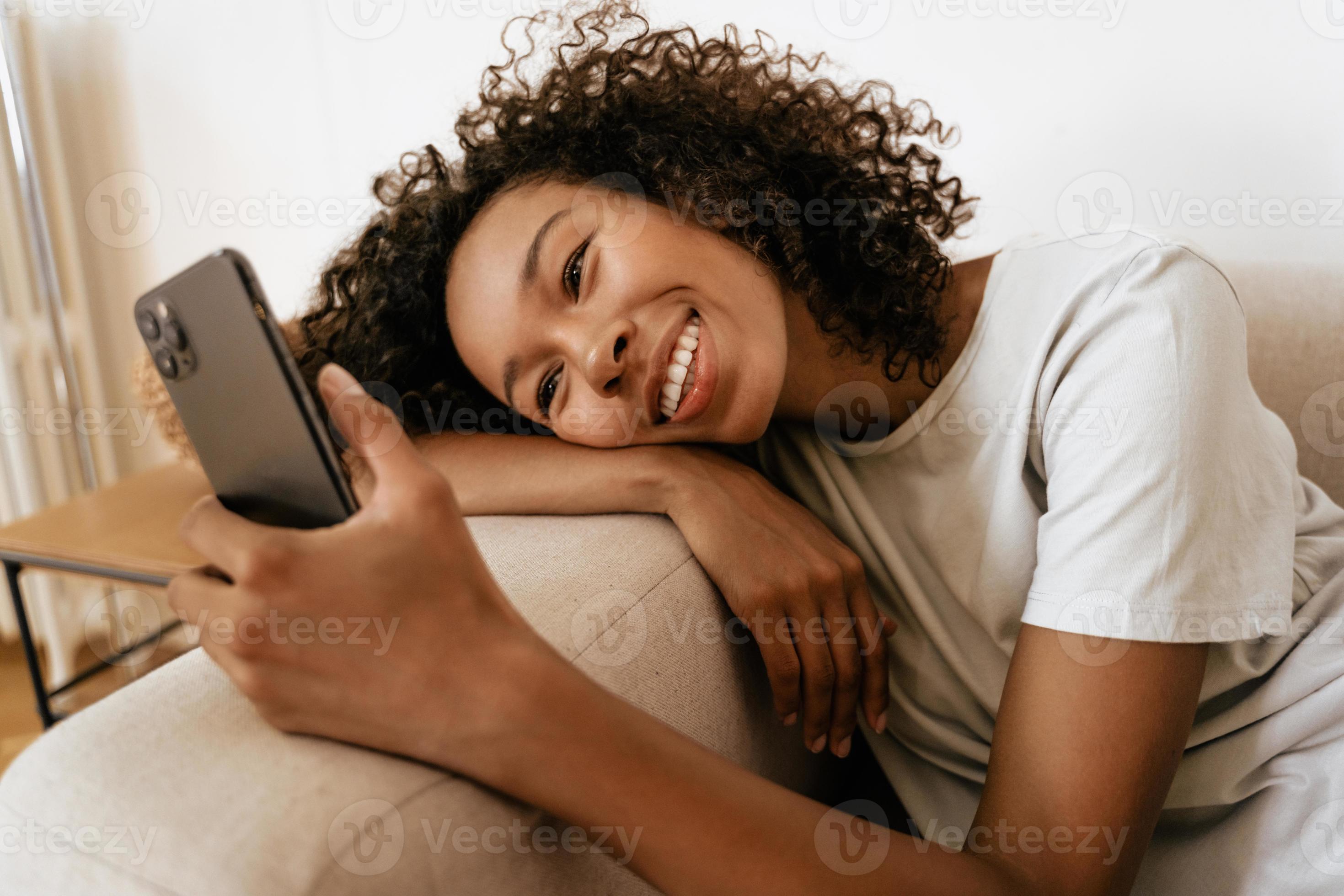 Black Young Woman Using Mobile Phone And Smiling While Resting On Sofa