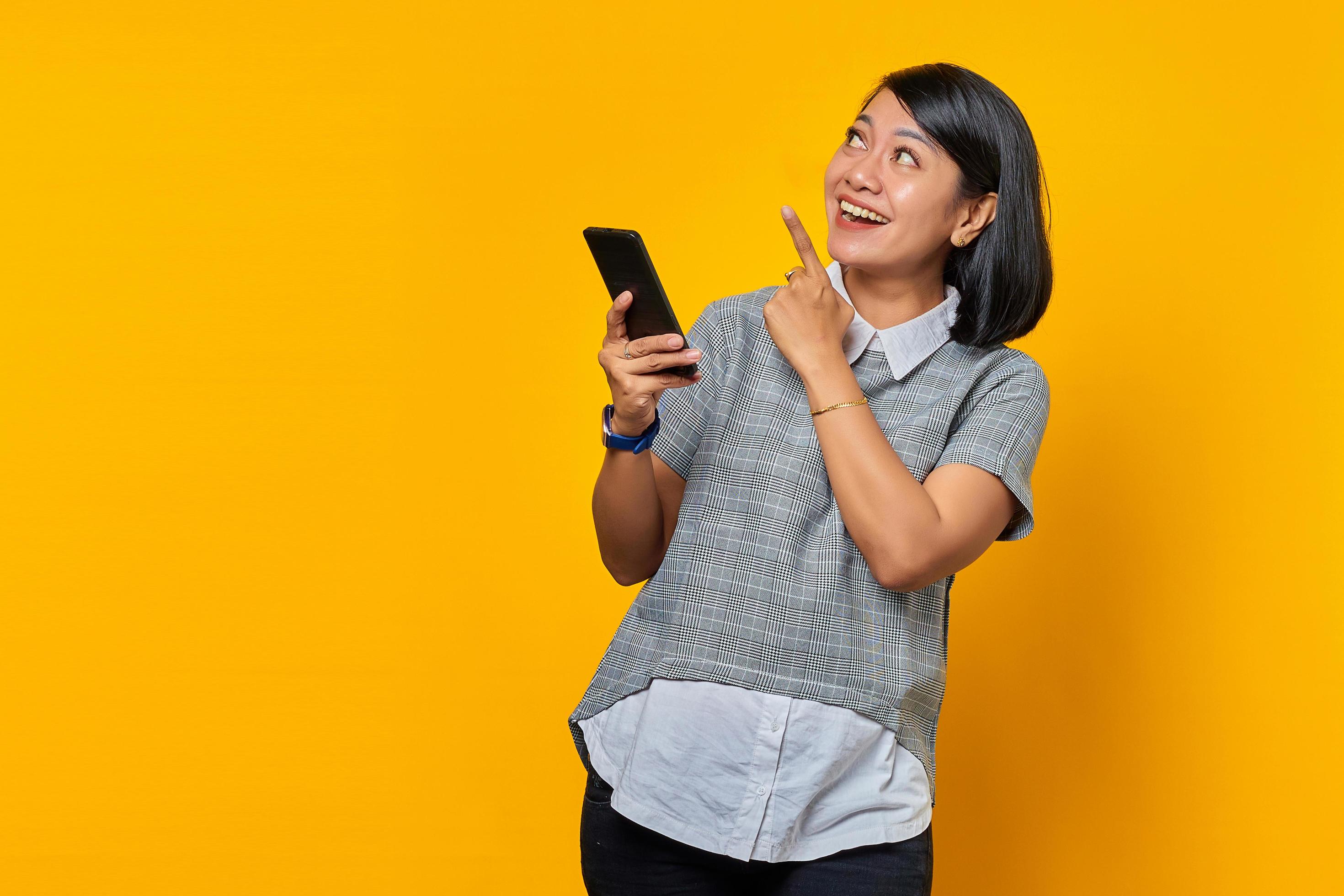 Cheerful Young Asian Woman Holding Mobile Phone And Pointing Finger Up