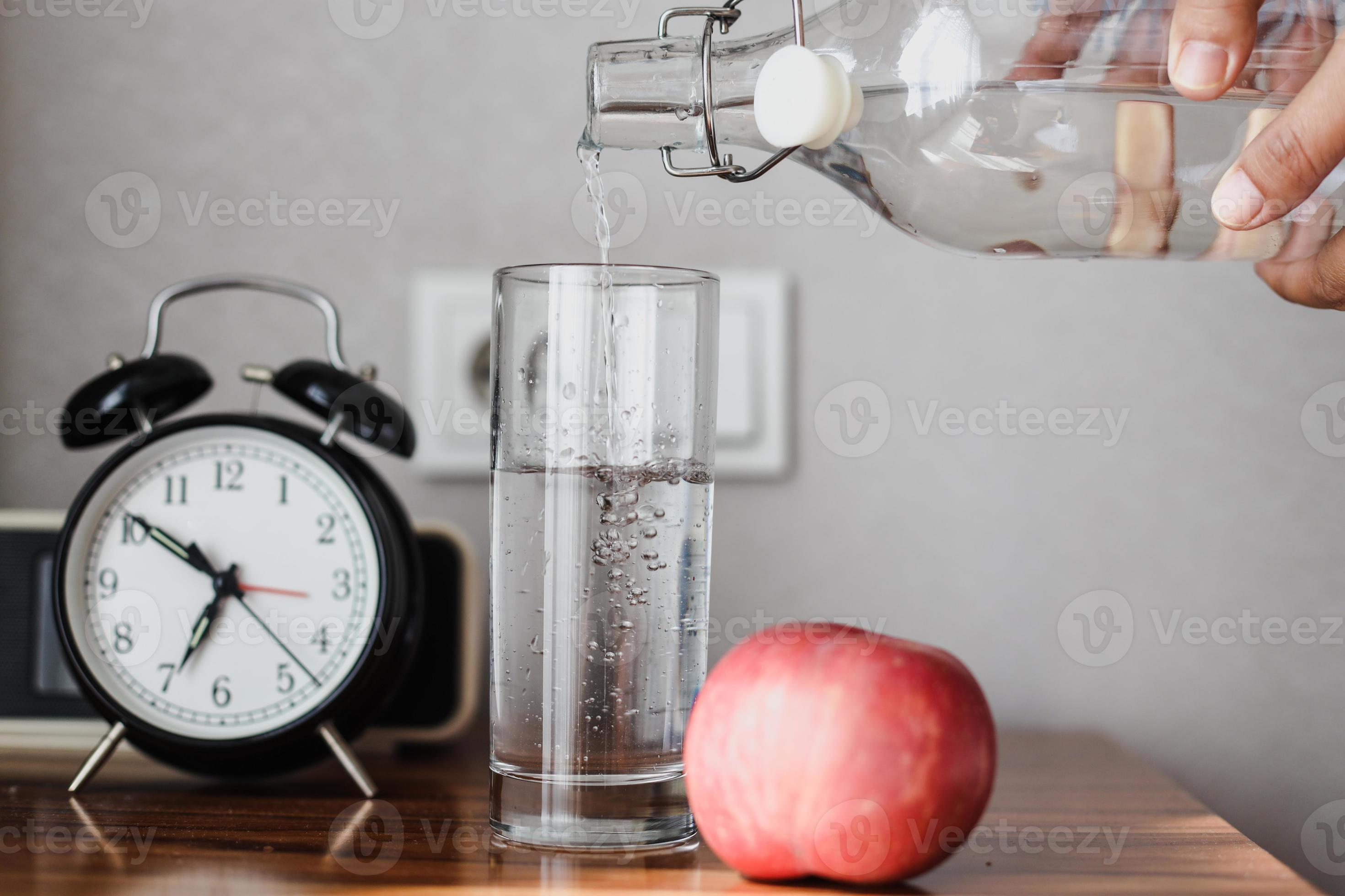 Hand pouring mineral water from a bottle to a glass with an apple on the table for breakfast ...