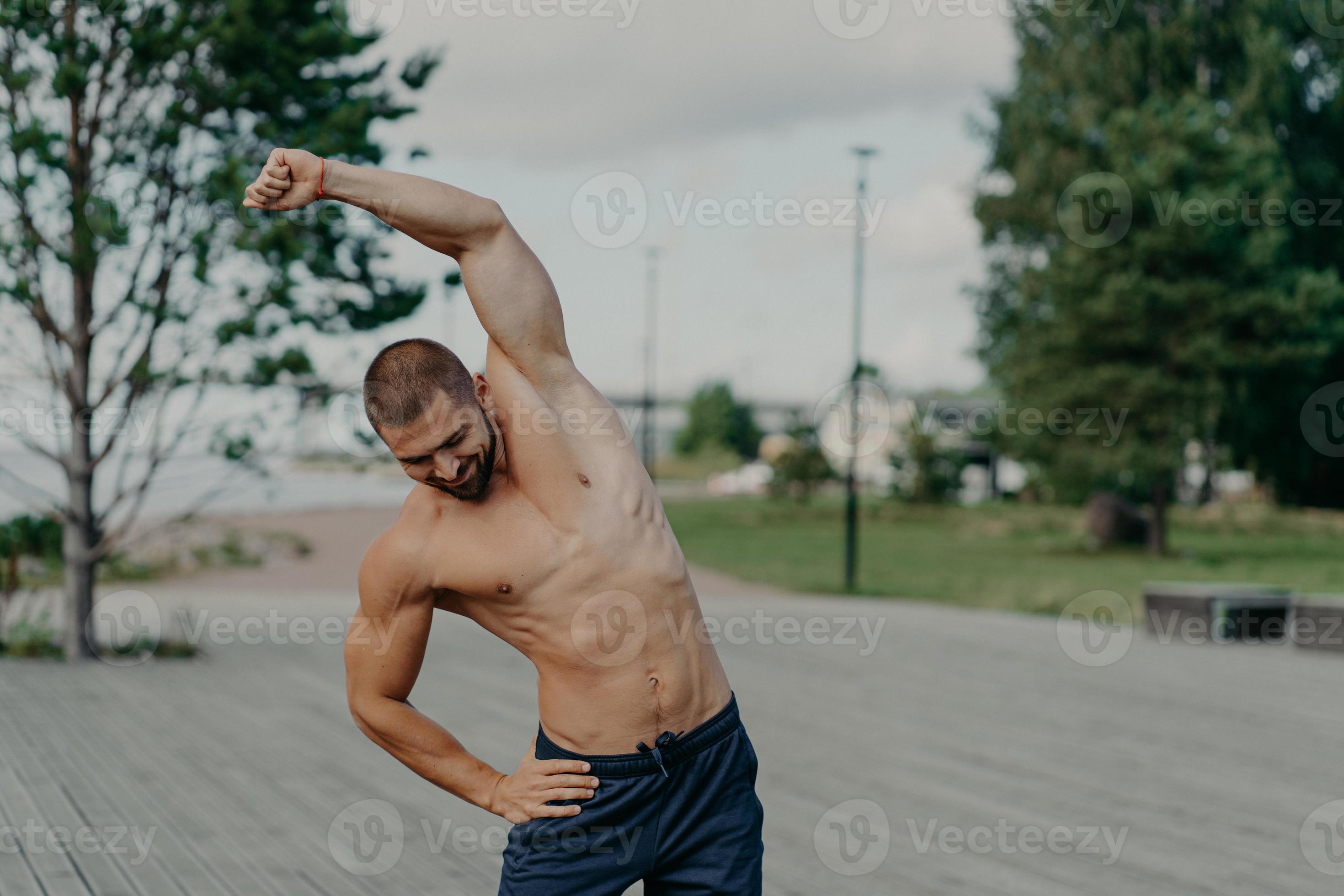 Outdoor shot of active muscular man makes stretching excerises and poses with naked torso ...