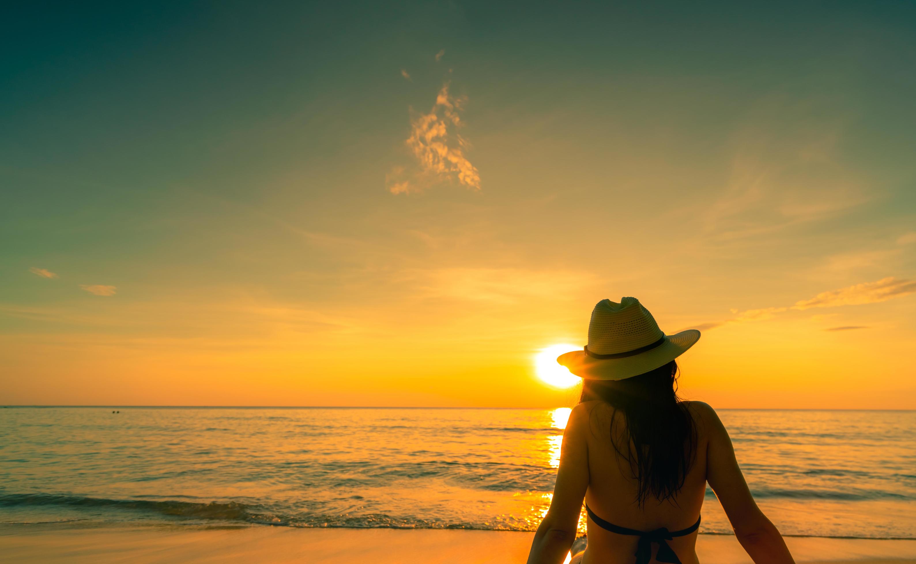 Back View Of Asian Woman Wear Bikini And Straw Hat Sit On Sand Beach