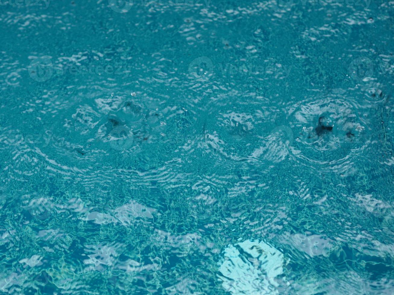 Rain drops falling blue water in pool background ripples on the surface texture, glittering ...