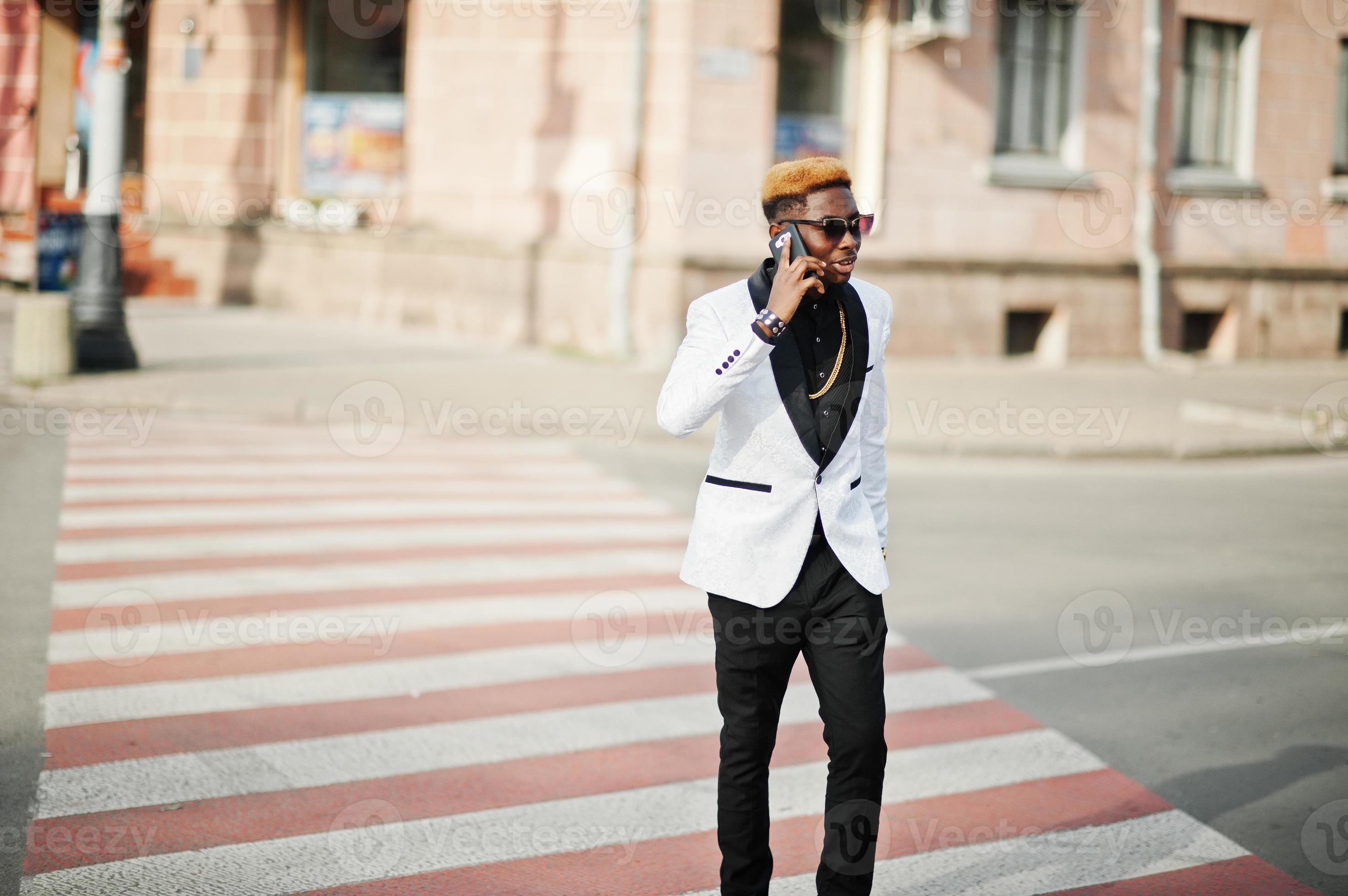 Chic handsome african american man in white suit walking on crosswalk and speaking on phone ...