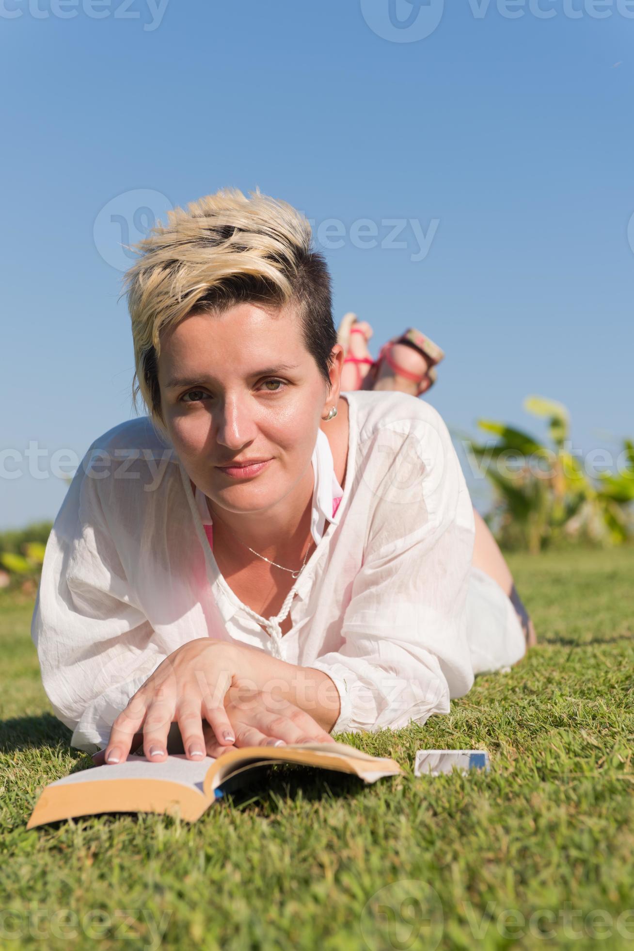 Woman Lying And Reading Her Favorite Book On A Meadow Covered With Fresh Green Grass On A Sunny