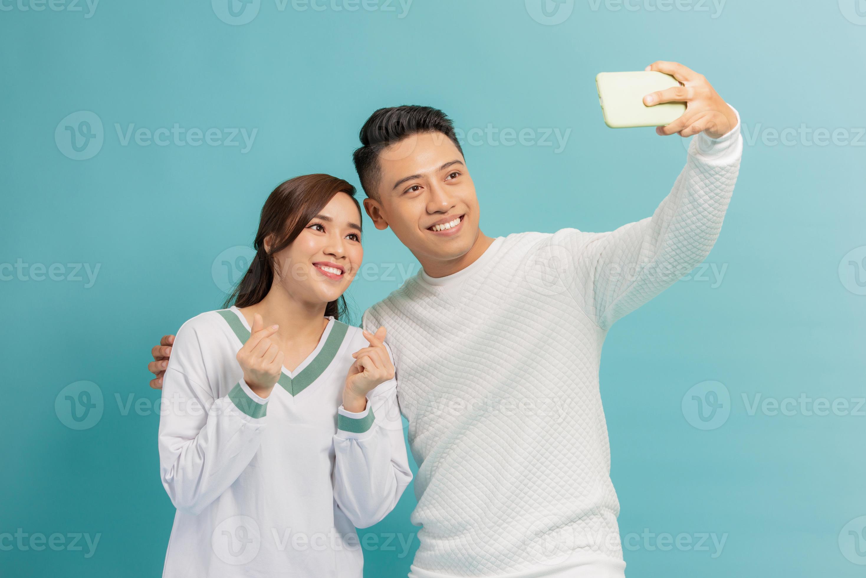 Happy young couple showing heart korean style while taking a selfie together isolated over blue ...