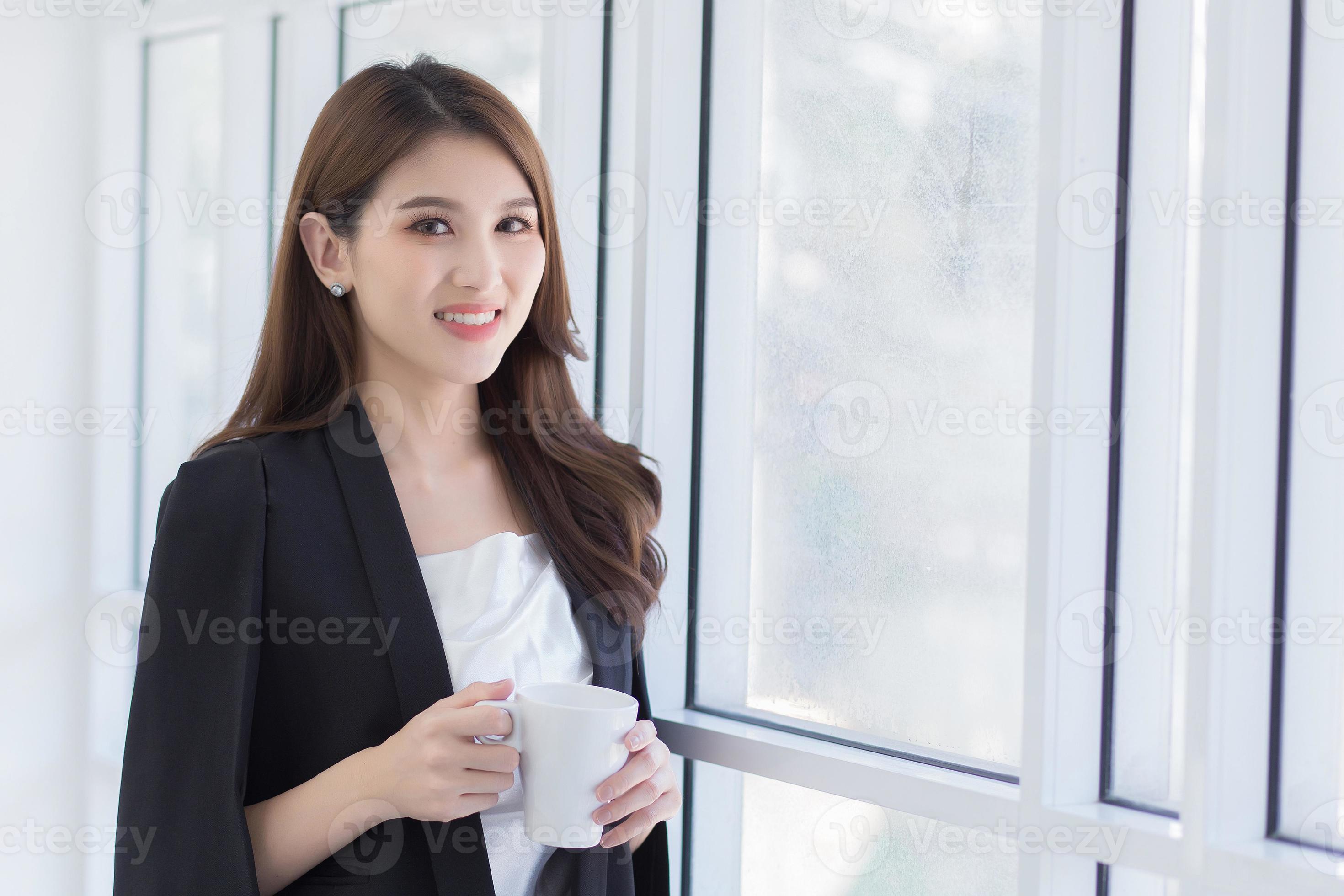 A beautiful office lady standing by holding a coffee cup by the window Happily in the workplace ...