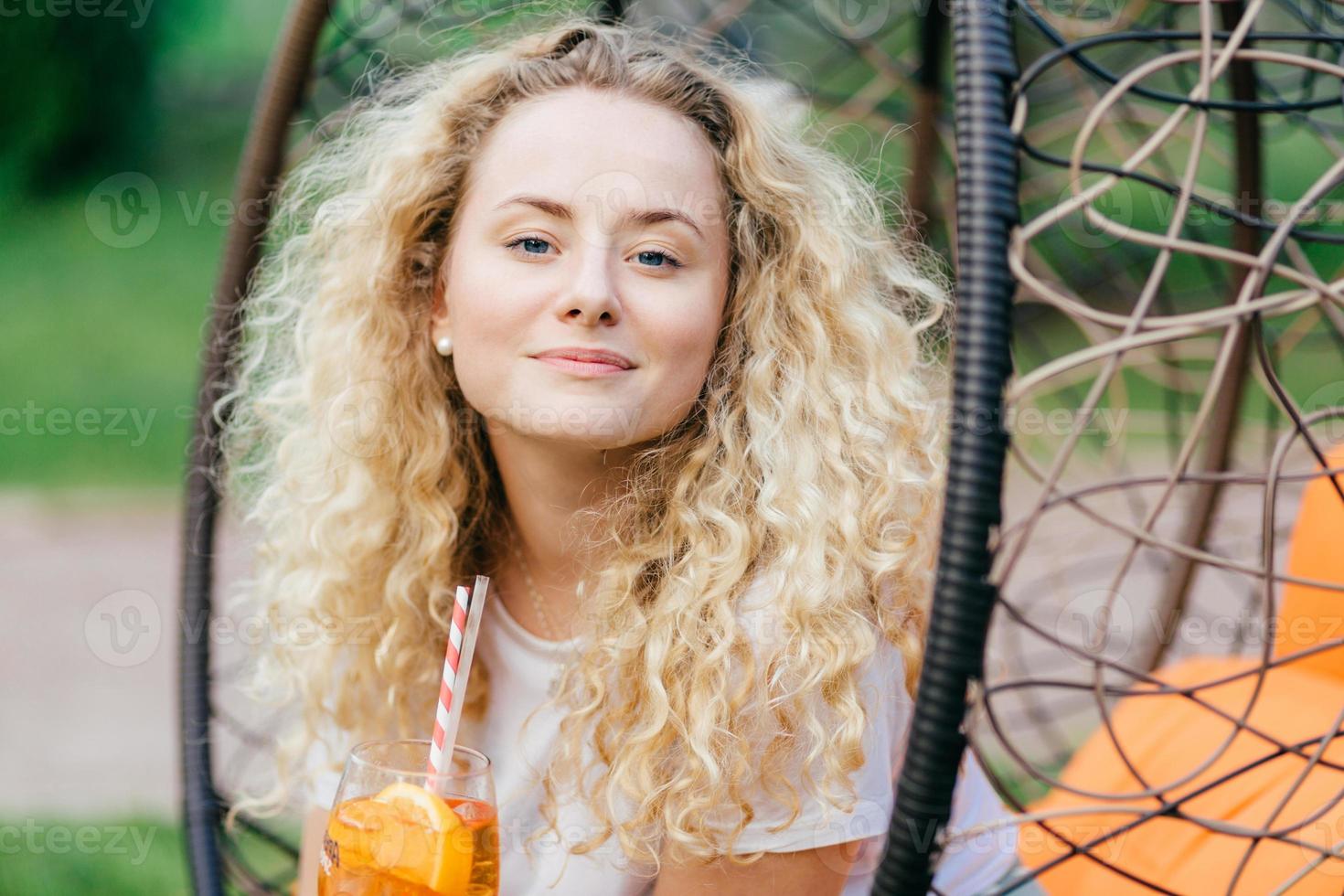 Confident Beautiful Female With Light Curly Hair Drinks Summer Cocktail Sits On Hanging Chair