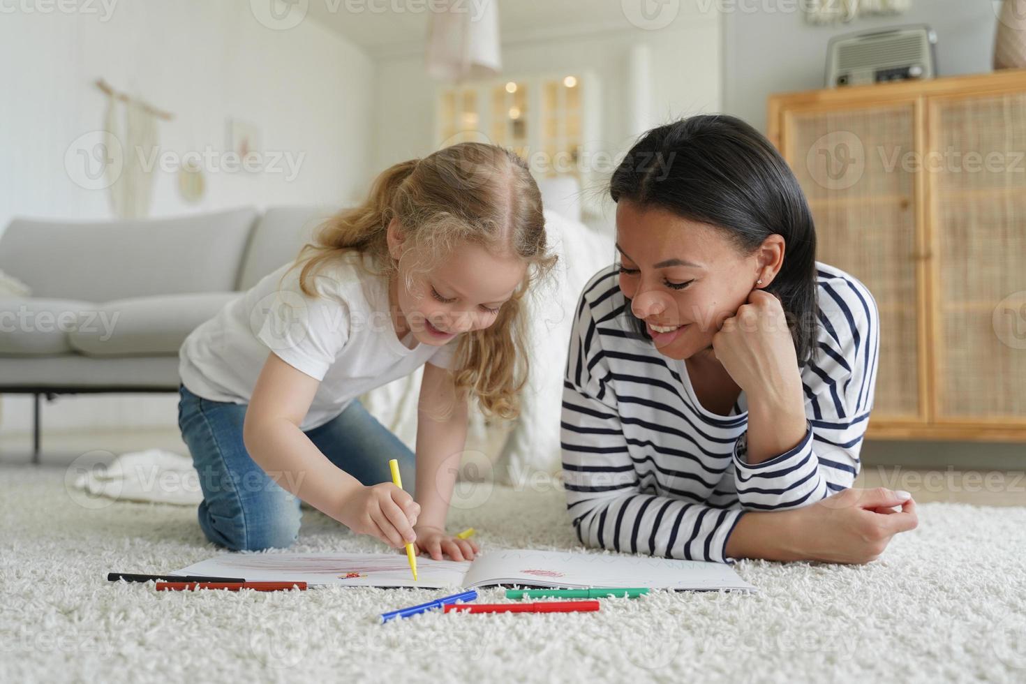 Mom, adopted child daughter draw with colorful pencils, lying on floor. Happy motherhood ...