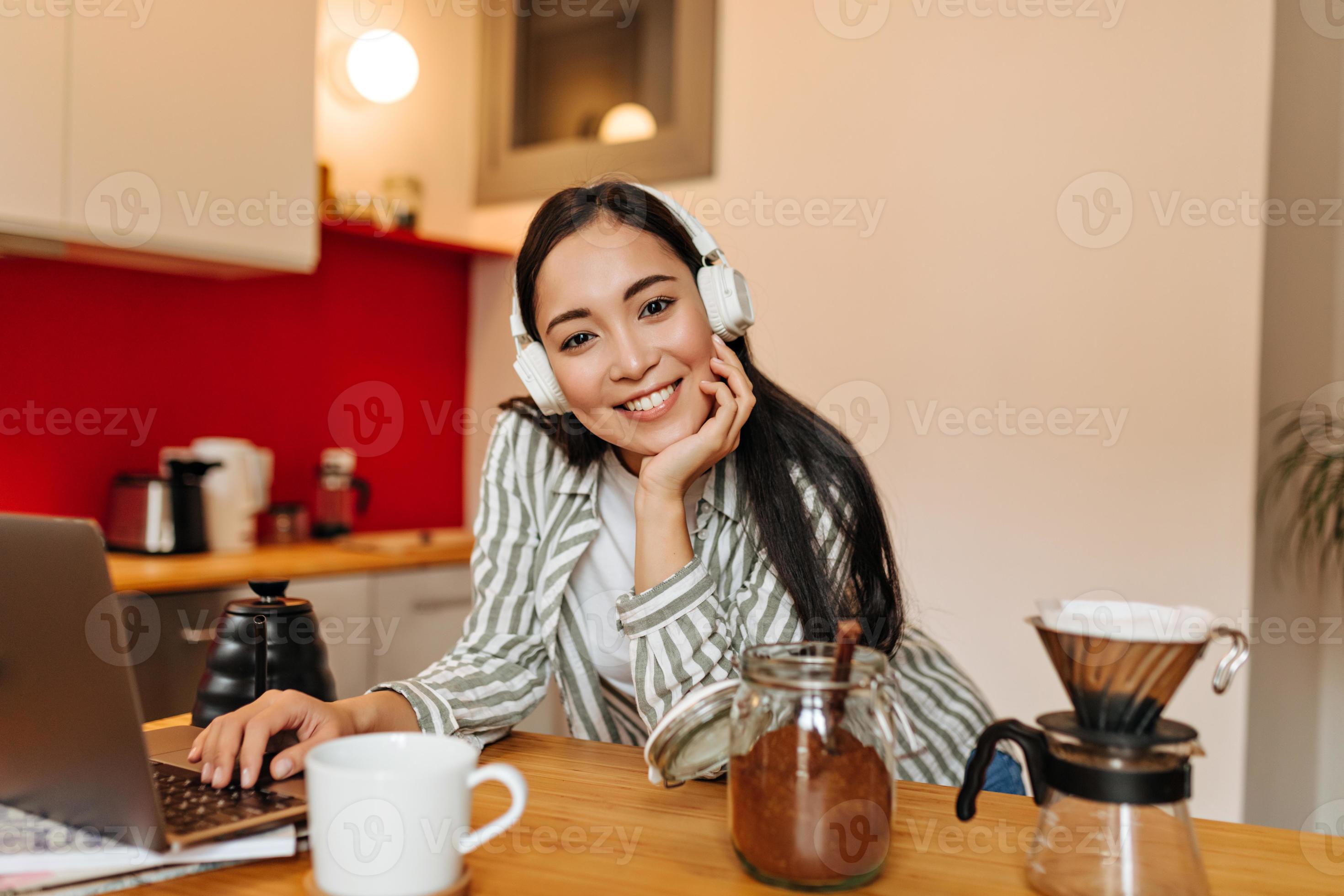 Brown-eyed girl leans on kitchen table, typing in laptop and listening to music on headphones ...