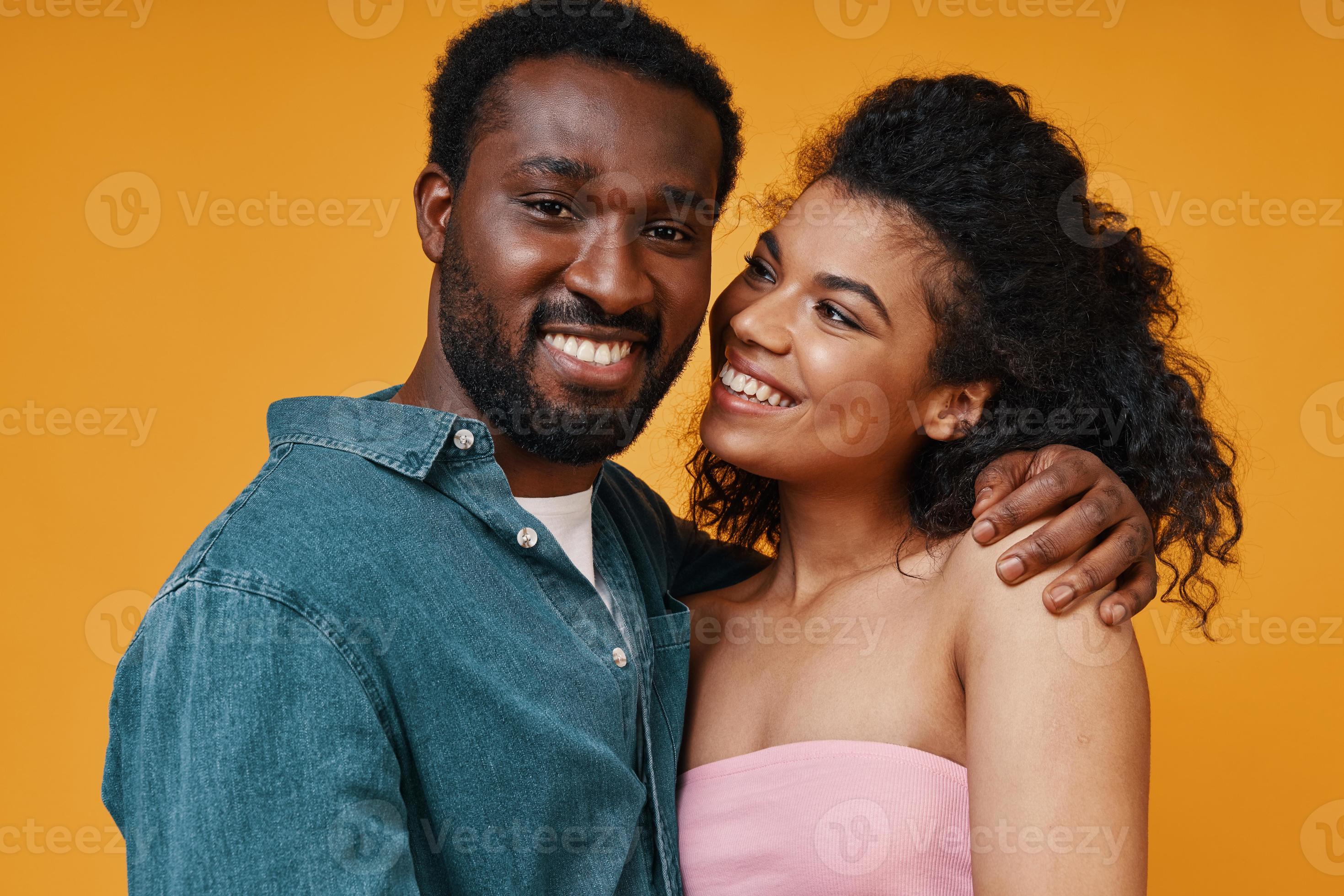 Beautiful young African couple hugging and smiling while standing against yellow background ...