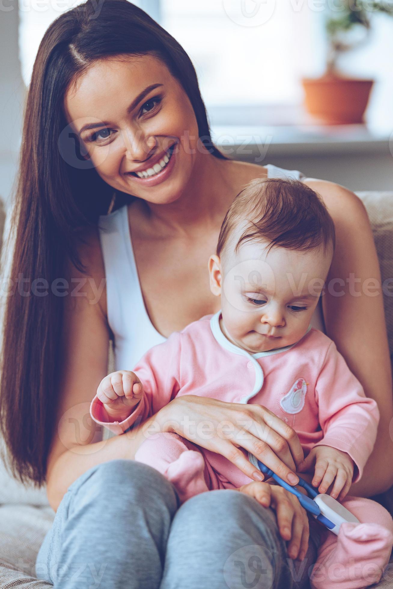 Best mom in the world. Beautiful young woman holding baby girl with toy on her knees and looking ...