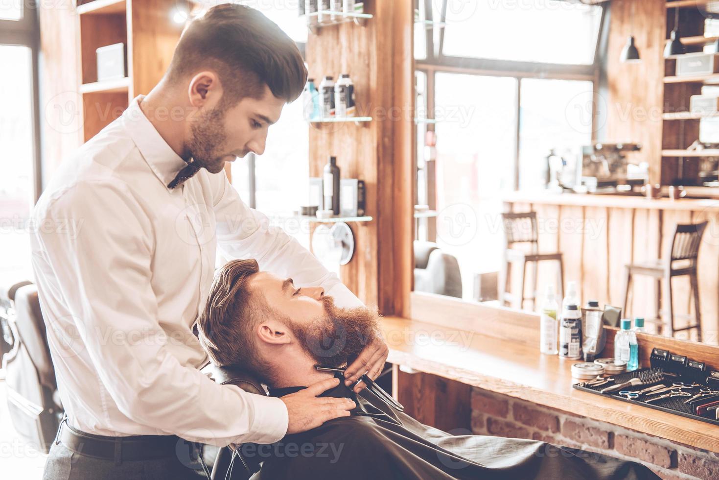 Making his beard stylish. Side view of young bearded man getting shaved ...