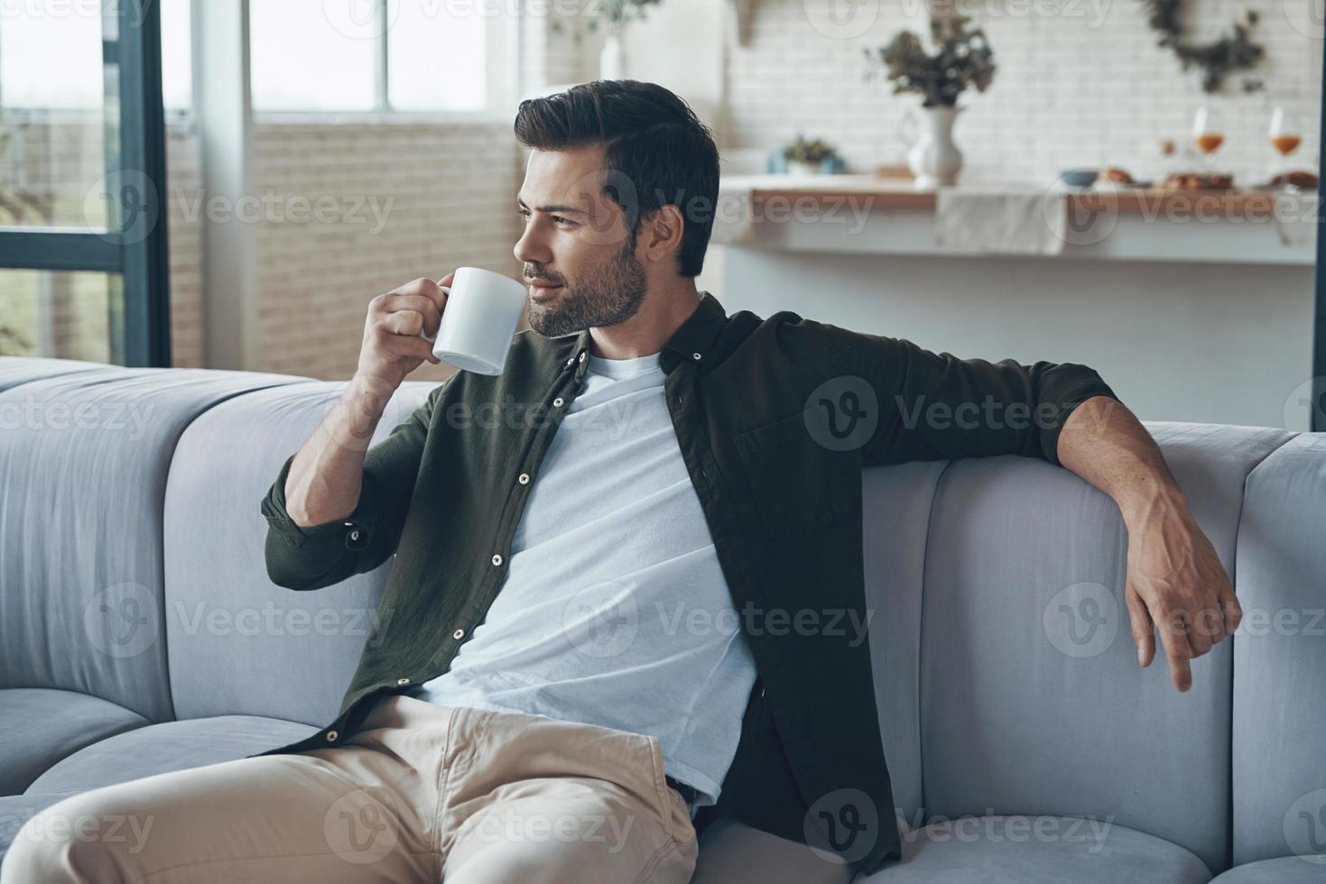 Thoughtful young man drinking coffee and looking away while sitting on ... Thoughtful young man drinking coffee and looking away while sitting on ...