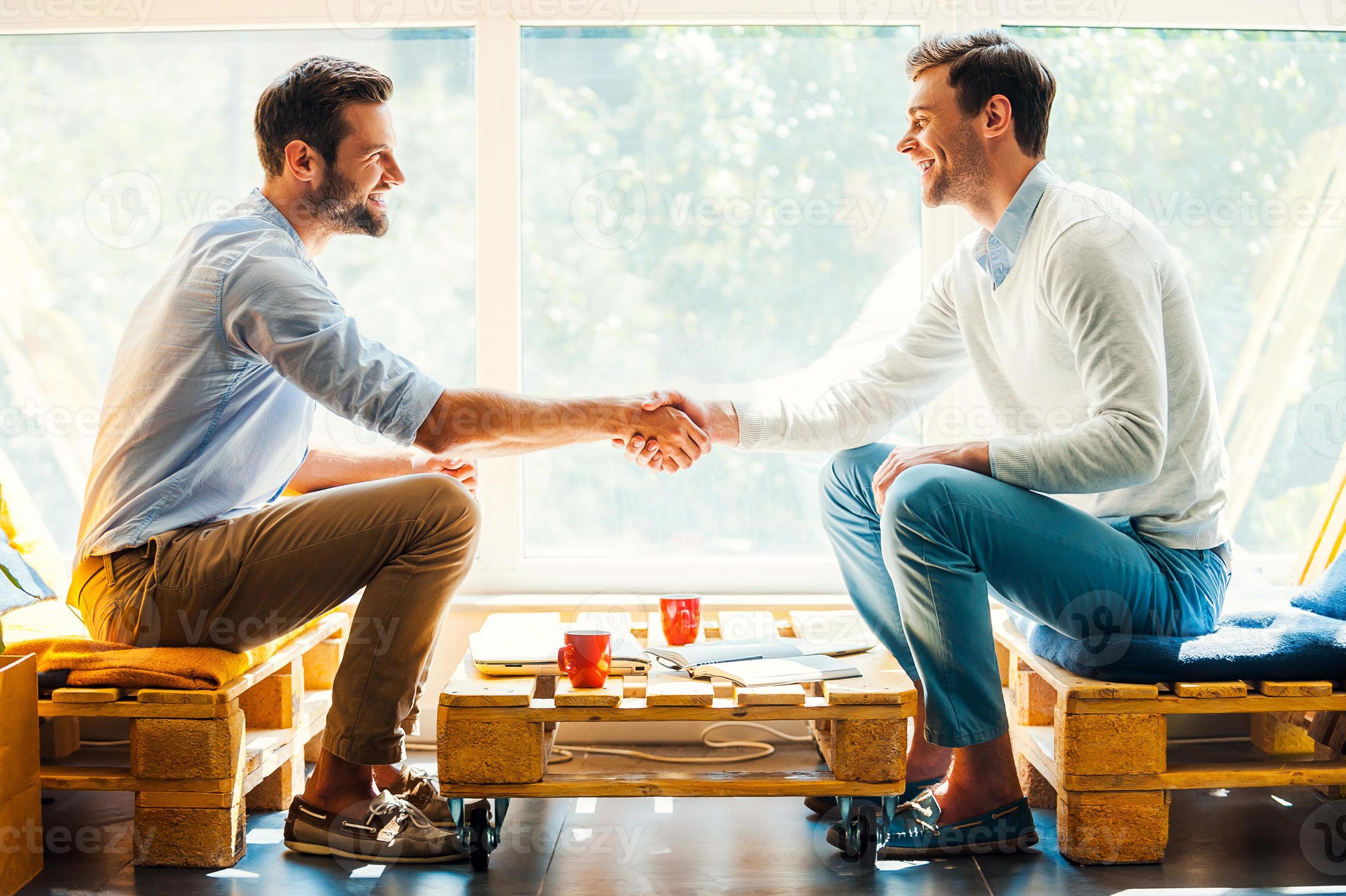 Men shaking hands. Side view of two happy young men shaking hands while ...