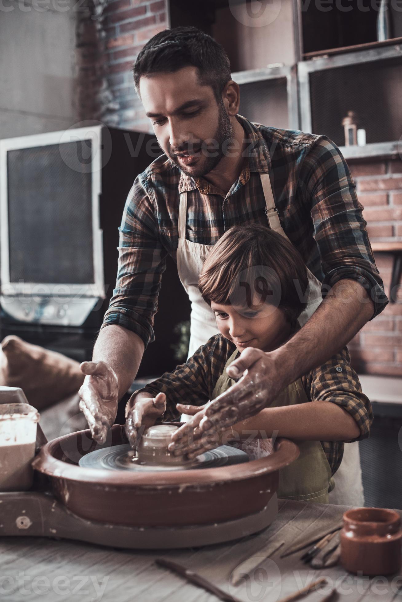 Pottering together. Confident young man and little boy making ceramic pot on the pottery class ...