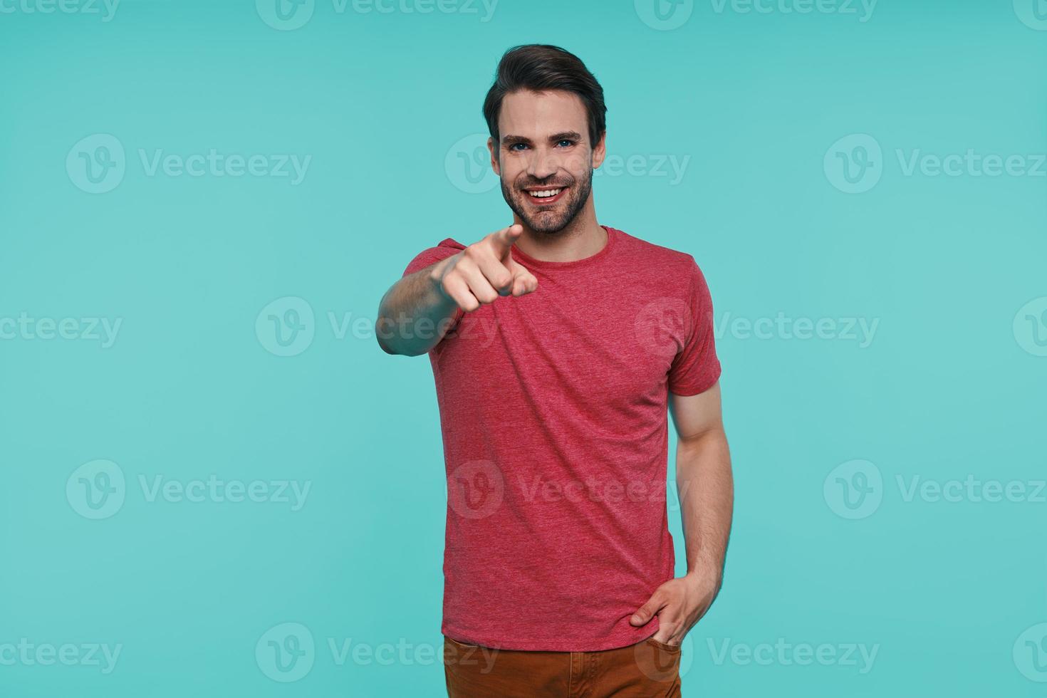 Handsome young smiling man pointing camera while standing against blue ...
