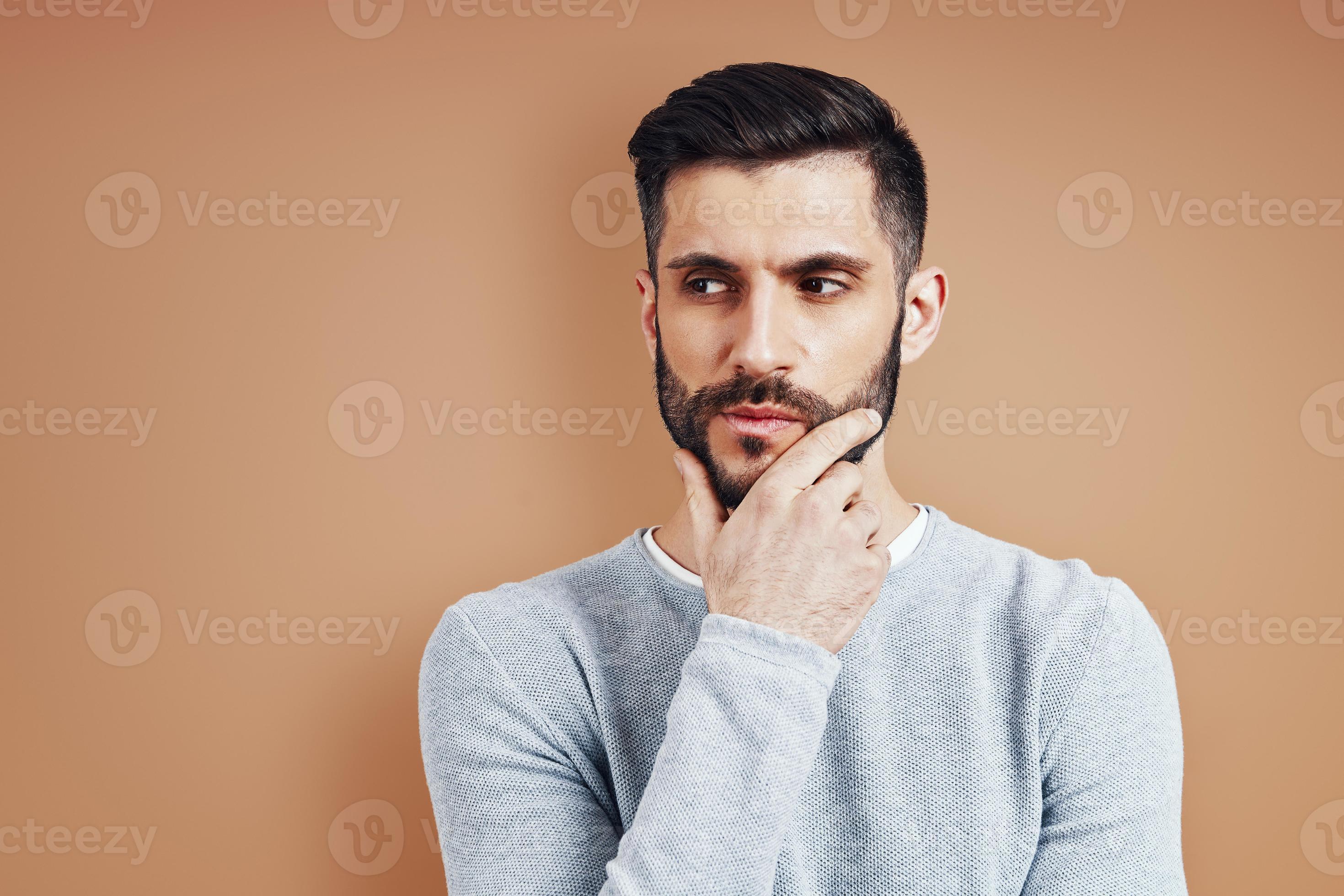 Thoughtful young man in smart casual wear keeping hand on chin while ...