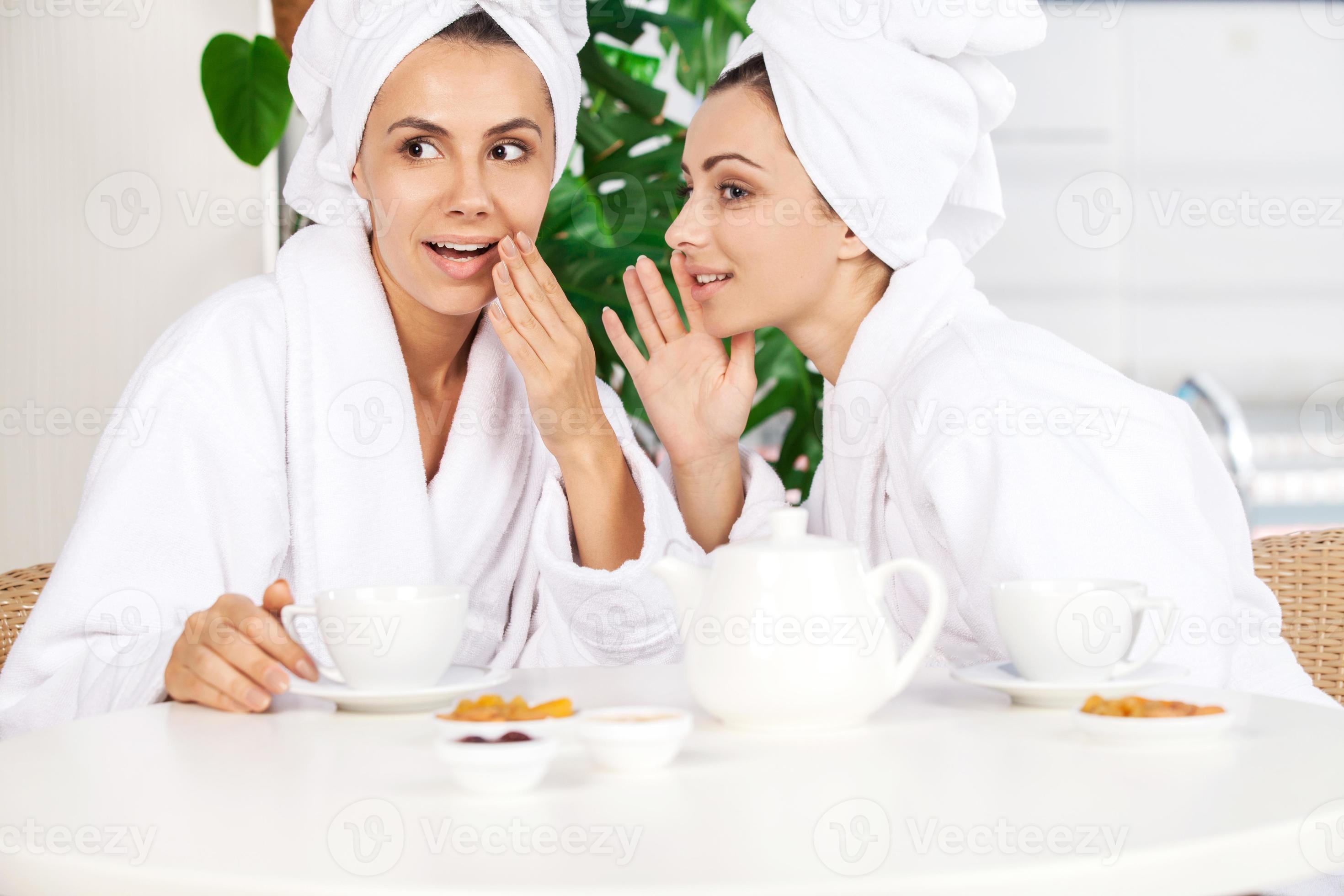 Girls at spa. Two beautiful young women in bathrobe drinking tea and gossiping while sitting in ...