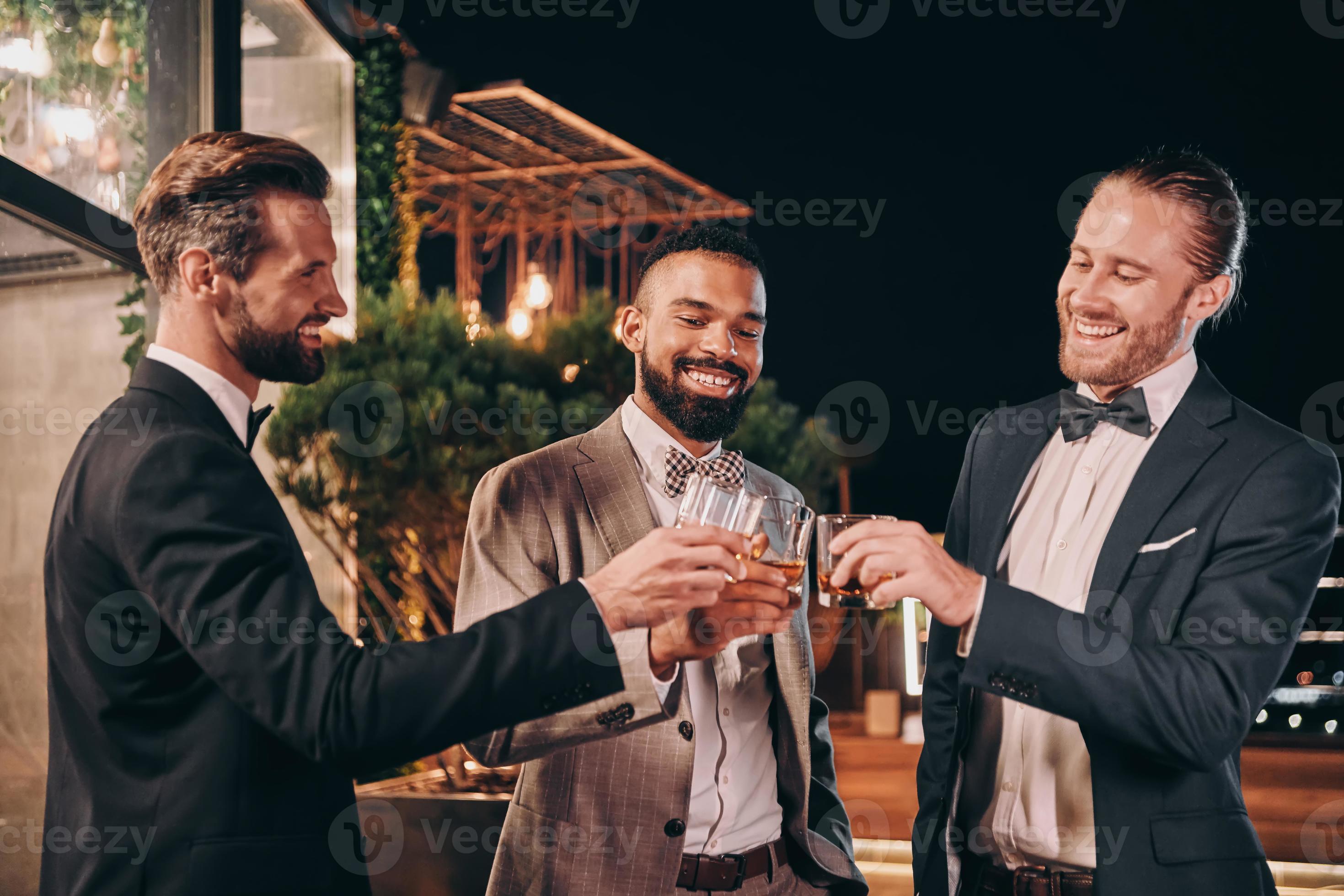 tres hombres guapos en trajes brindando con whisky y sonriendo mientras pasan el tiempo en la ...