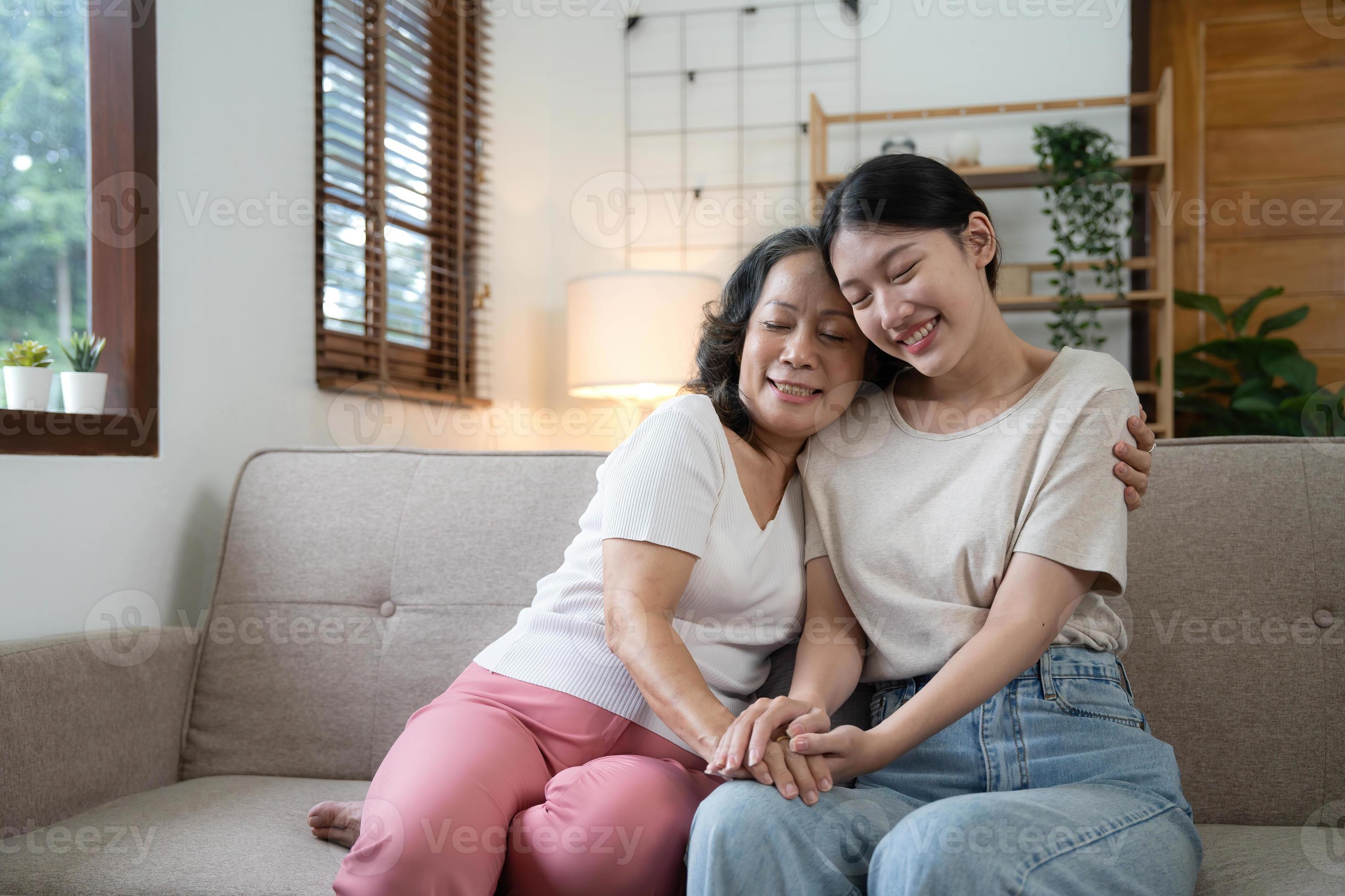Portrait happy adult daughter and older mother hugging and holding hands, sitting on couch at ...