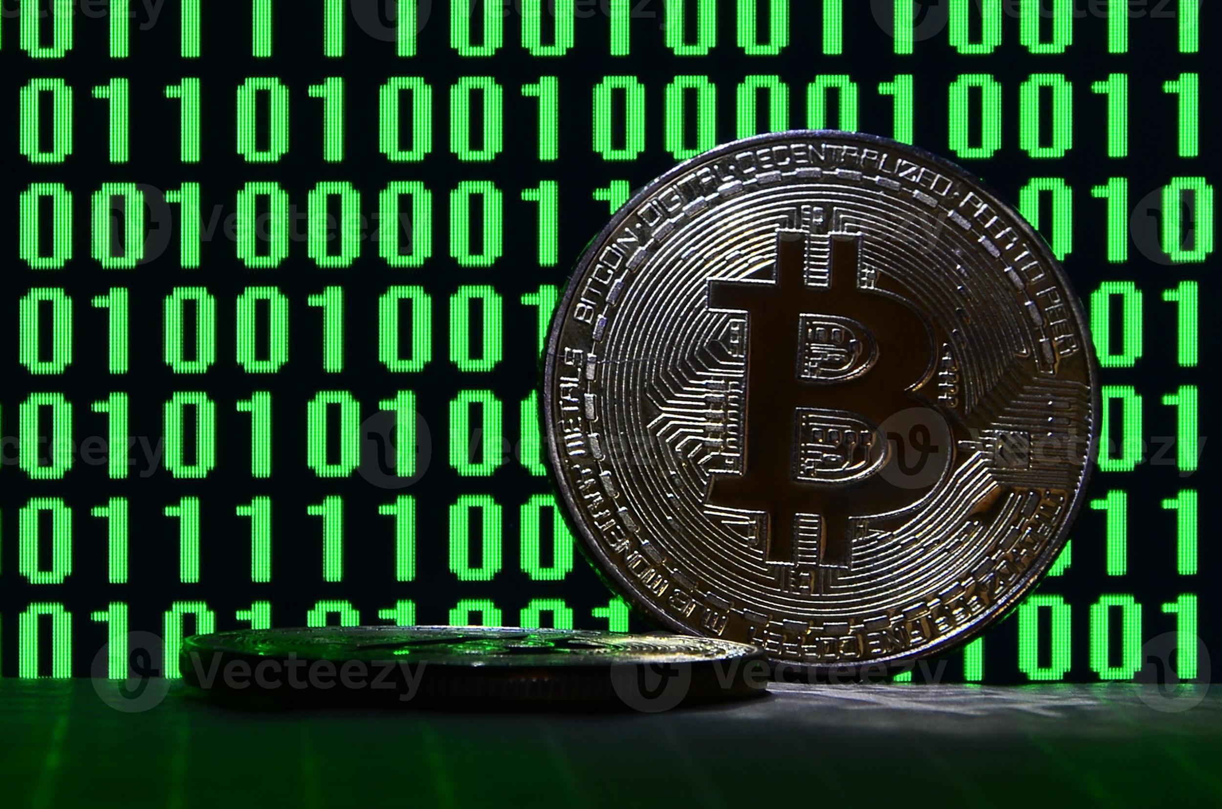 A pair of bitcoins lies on a cardboard surface on the background of a monitor depicting a binary ...
