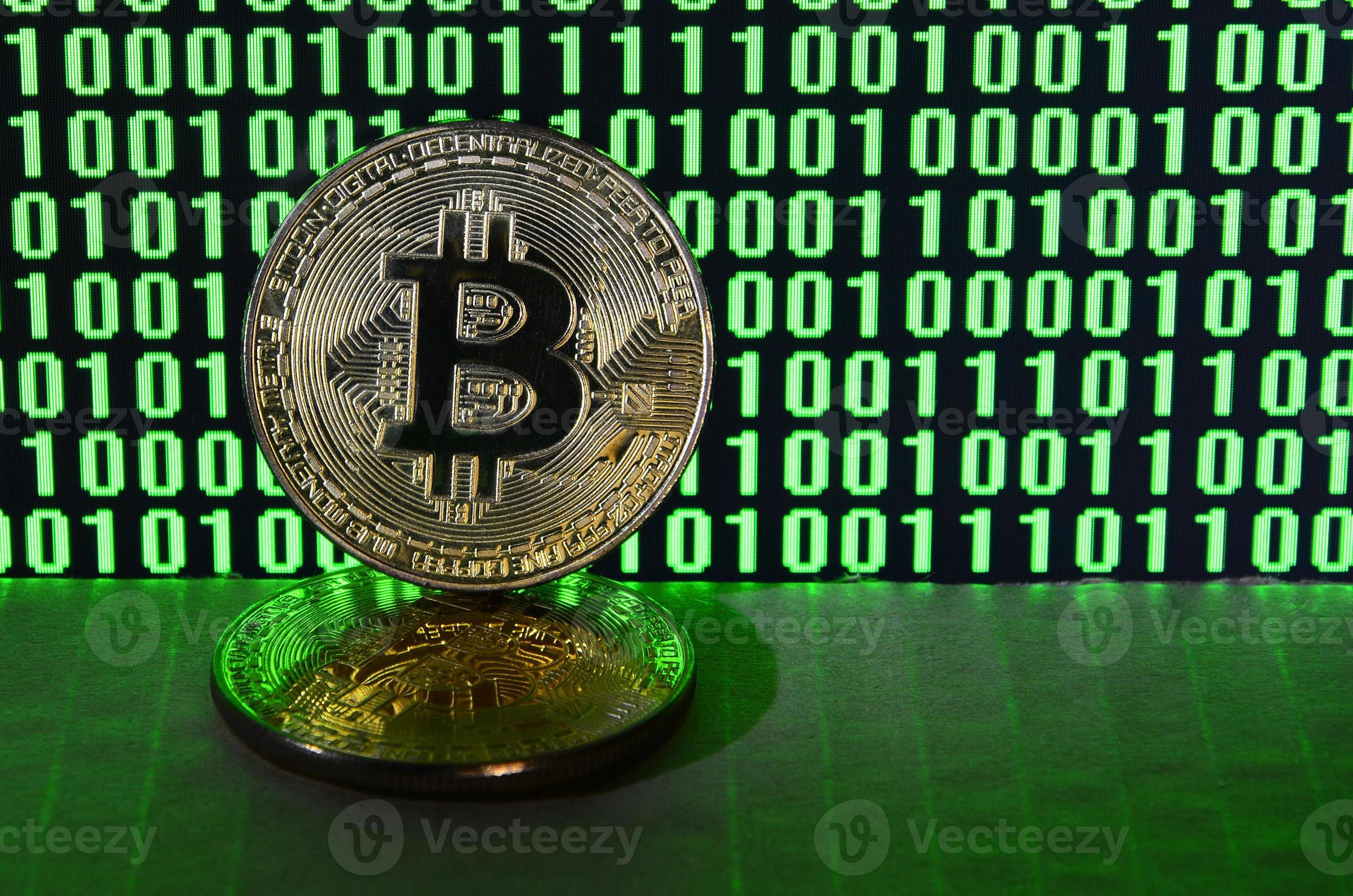 A pair of bitcoins lies on a cardboard surface on the background of a monitor depicting a binary ...