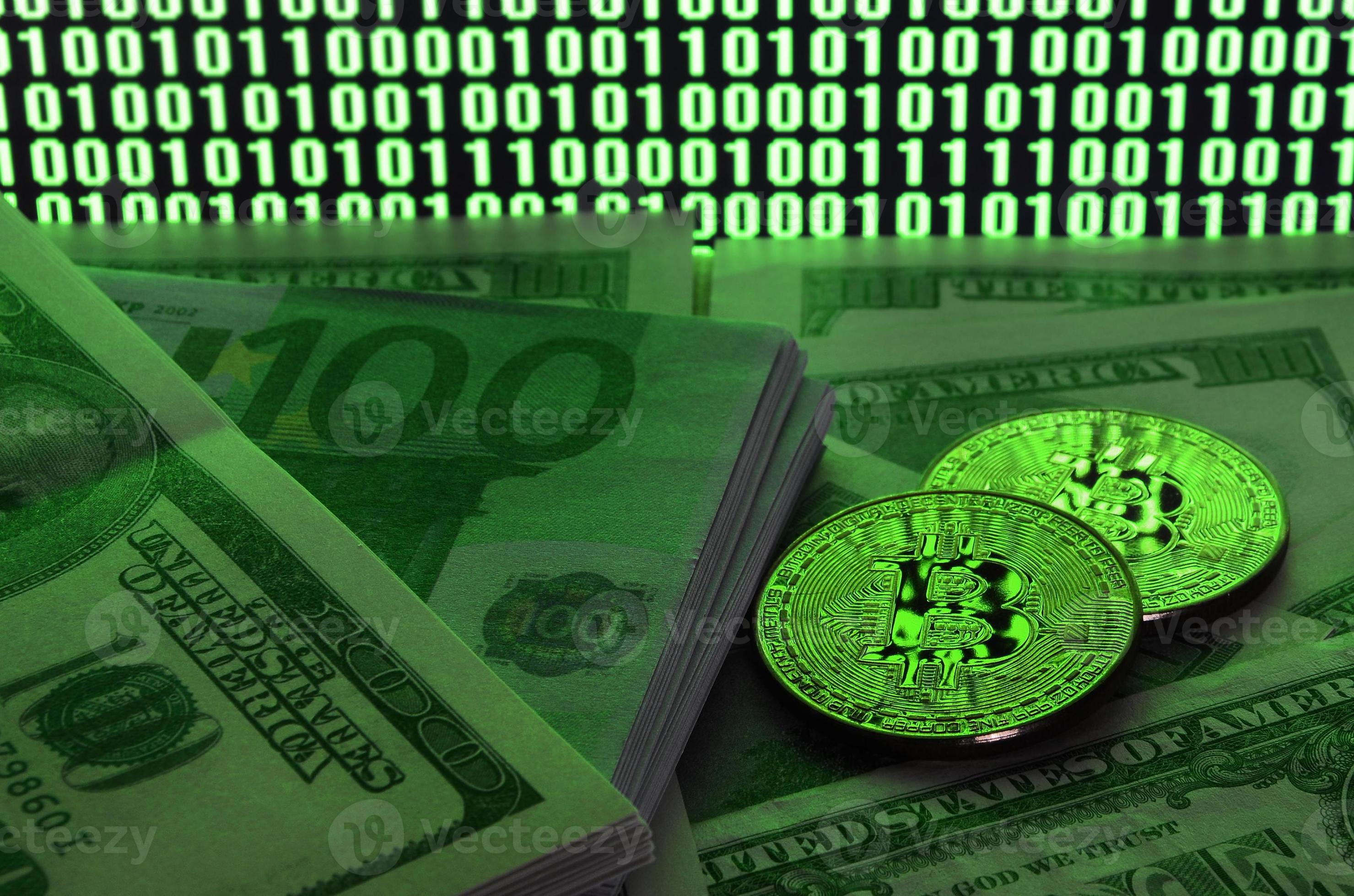 Two bitcoins lies on a pile of dollar bills on the background of a monitor depicting a binary ...