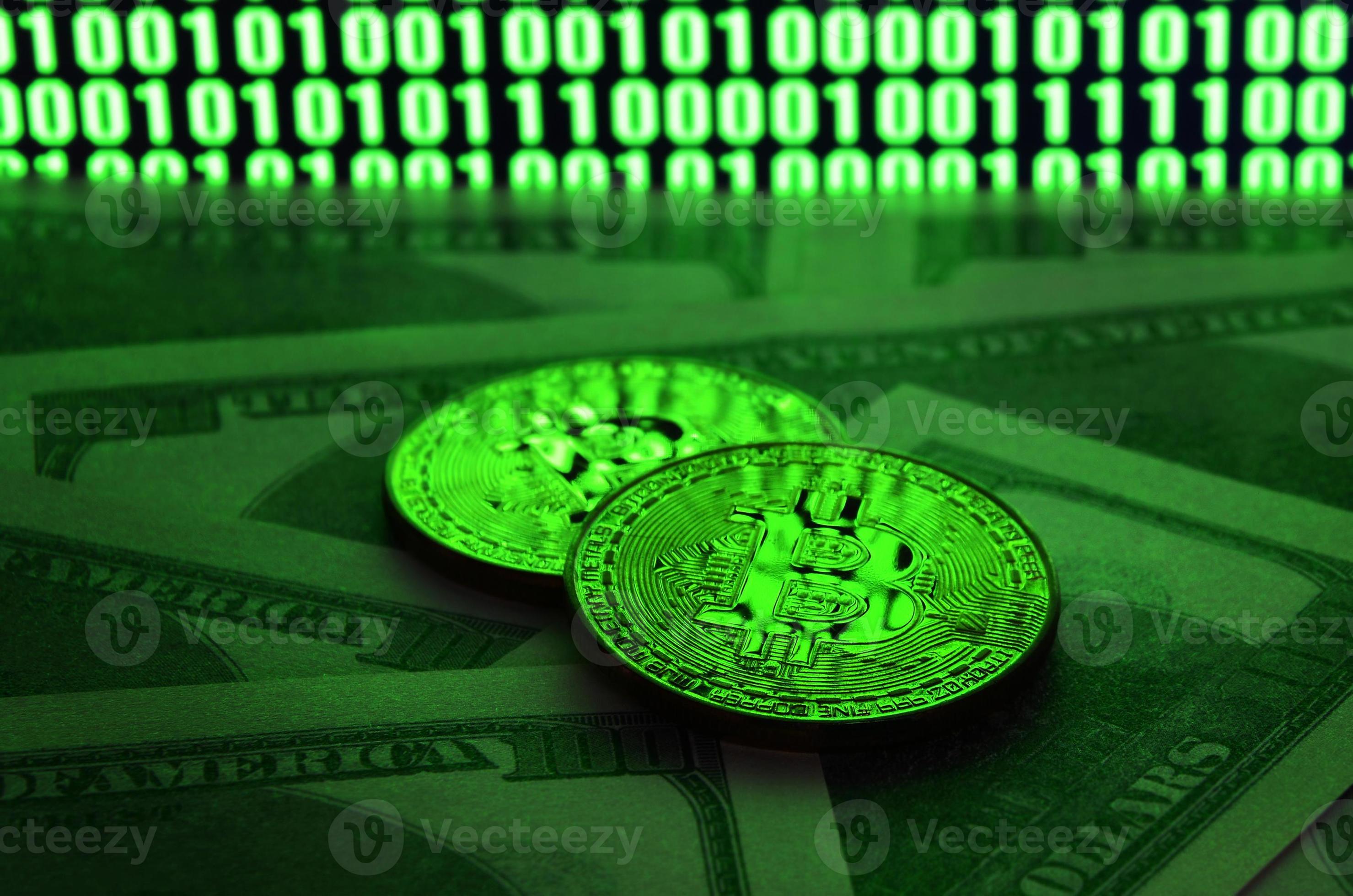 Two bitcoins lies on a pile of dollar bills on the background of a monitor depicting a binary ...