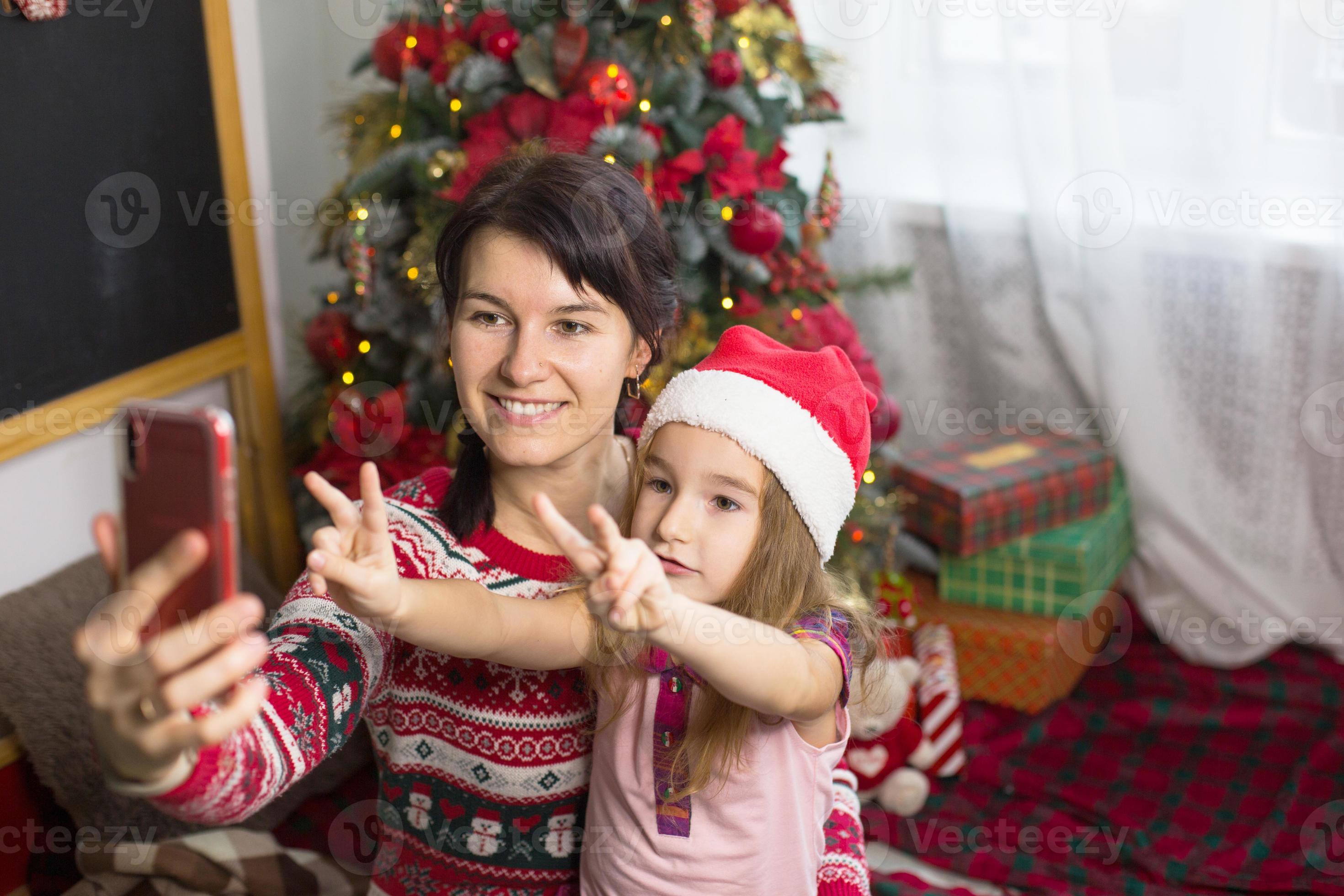 mamá e hija están sentadas cerca del árbol de navidad, abrazándose, tomando fotos y selfies por ...