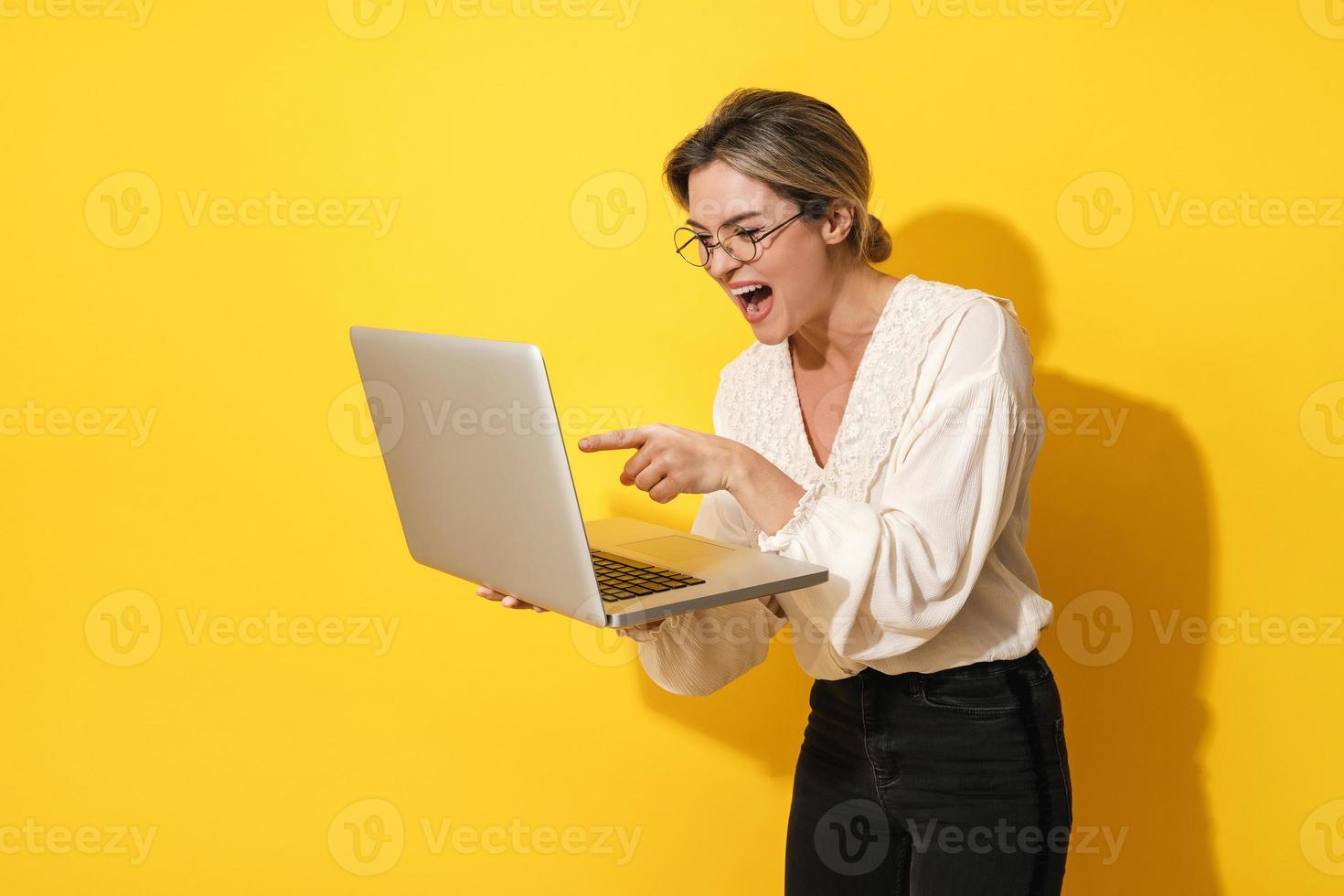 Cheerful woman wearing eyeglasses is using laptop computer on yellow ...