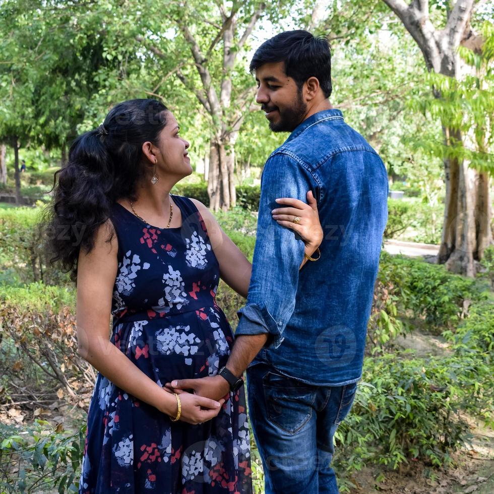 Indian couple posing for maternity baby shoot. The couple is posing in a lawn with green grass ...