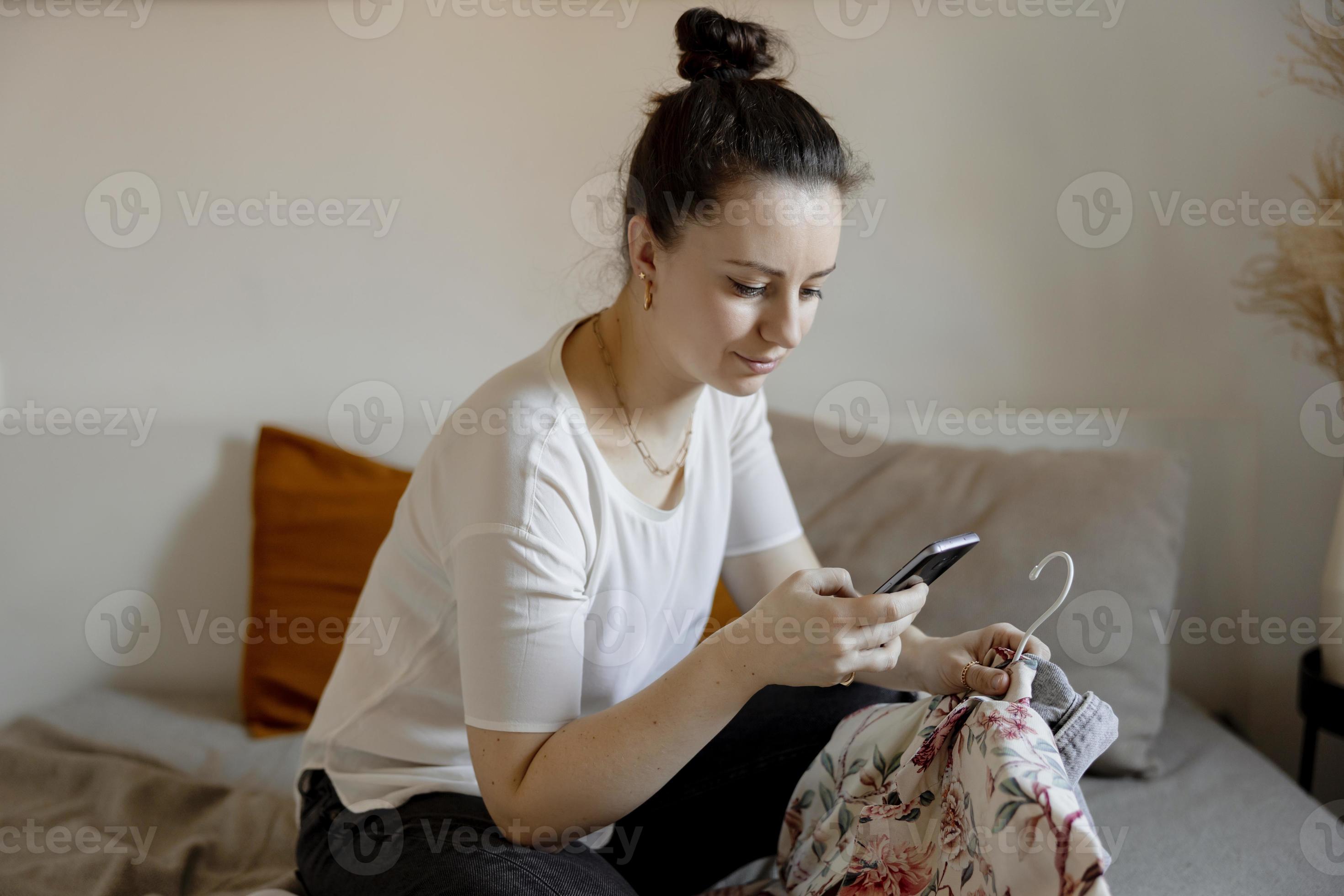 Young woman sitting on the bed at home and taking photo of her old clothes to sell them online ...