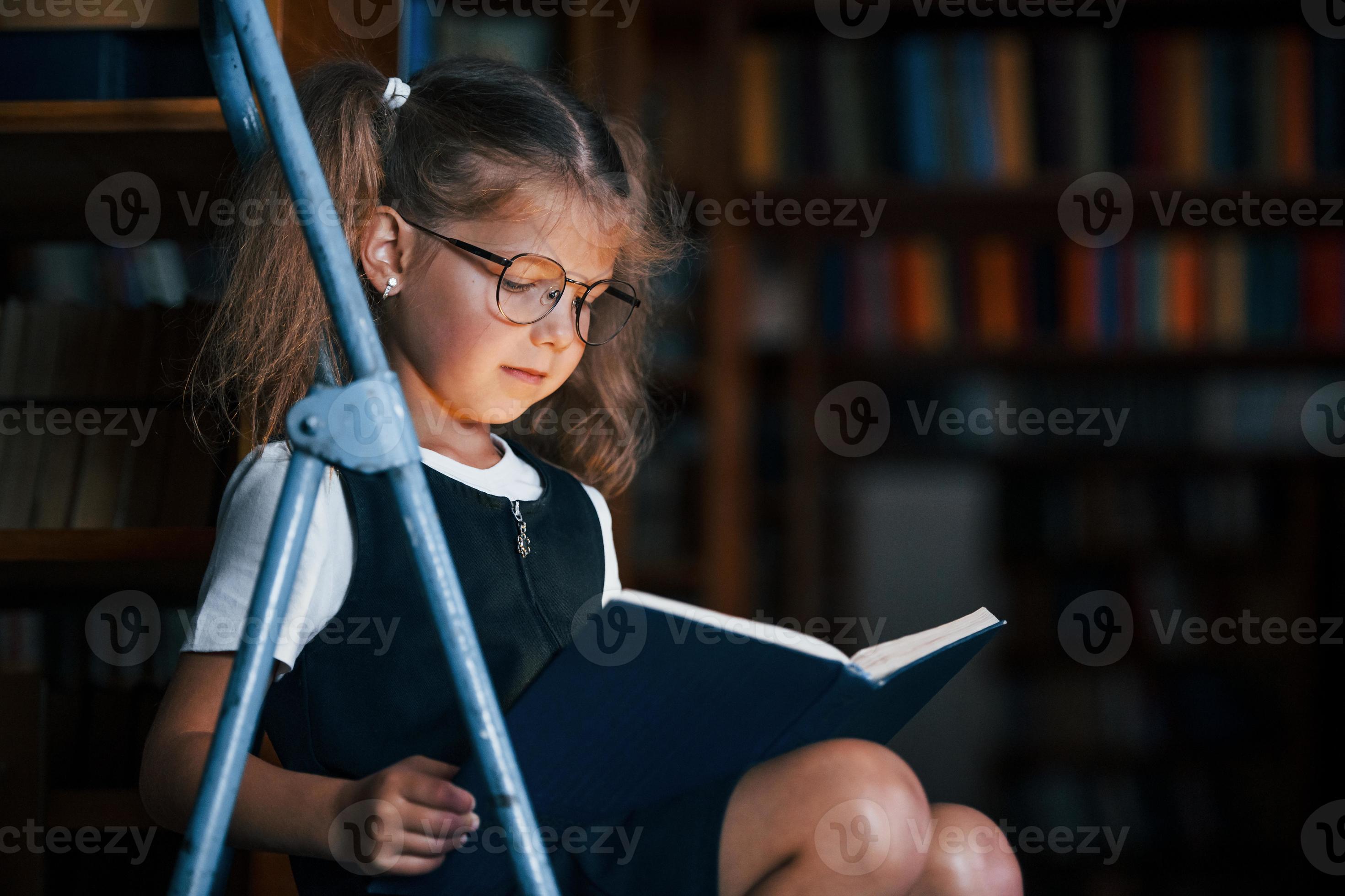 Concentration on reading. School girl on the ladder in library full of books. Education ...