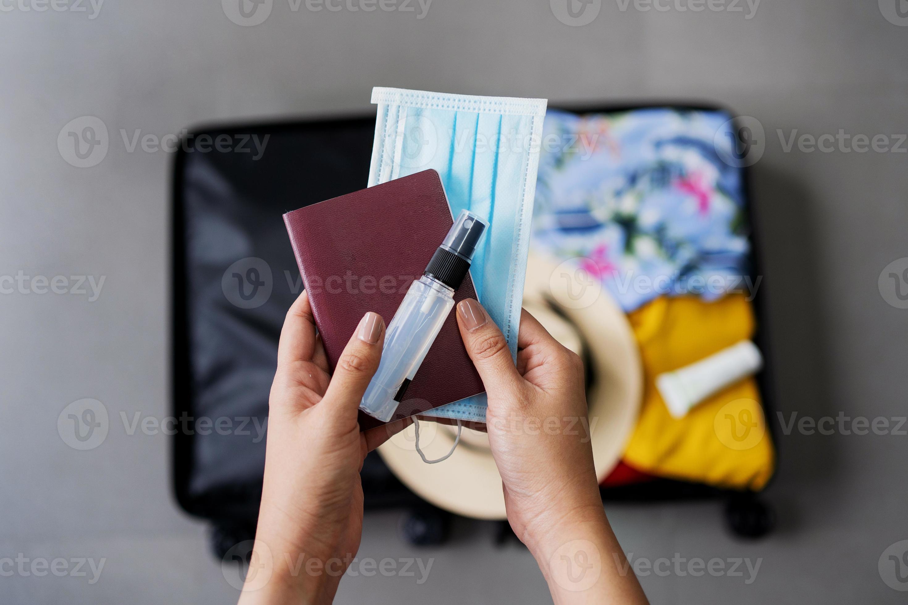 Young Woman Holding Passport Face Mask And Antiseptic Alcohol Spray On Suitcase Background