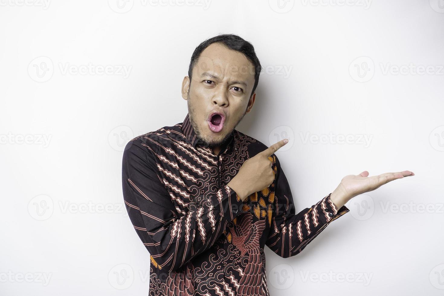 Shocked Asian man wearing batik shirt pointing at the copy space beside ...
