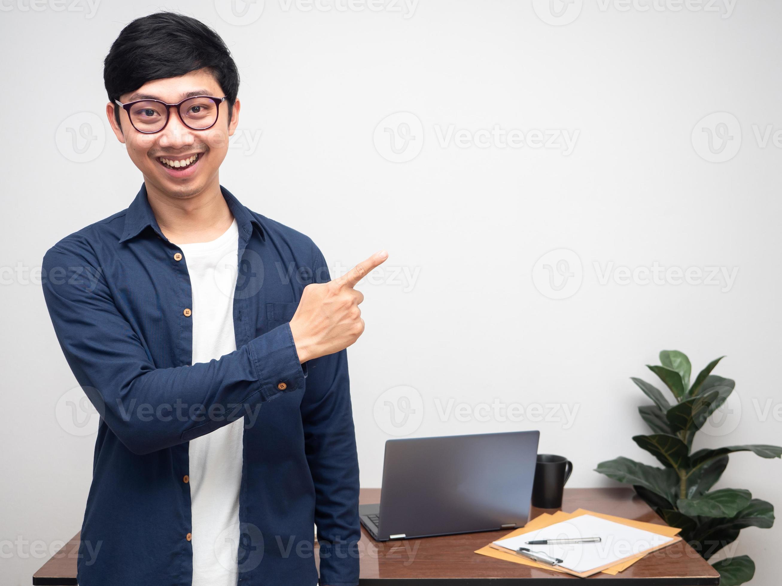 Positive businessman glasses smile stand cross arm at workplace room point finger at copy space ...