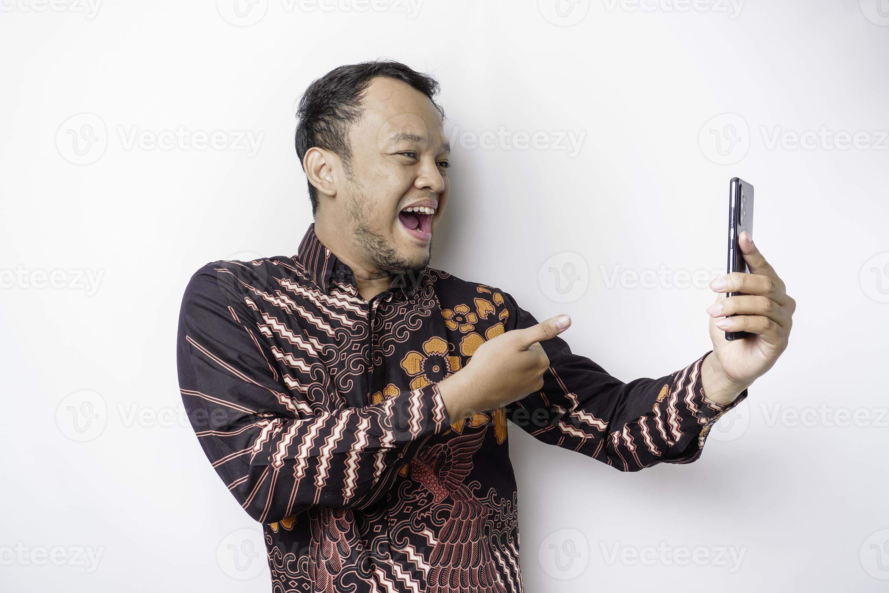 Shocked Asian man wearing batik shirt and holding his phone, isolated ...
