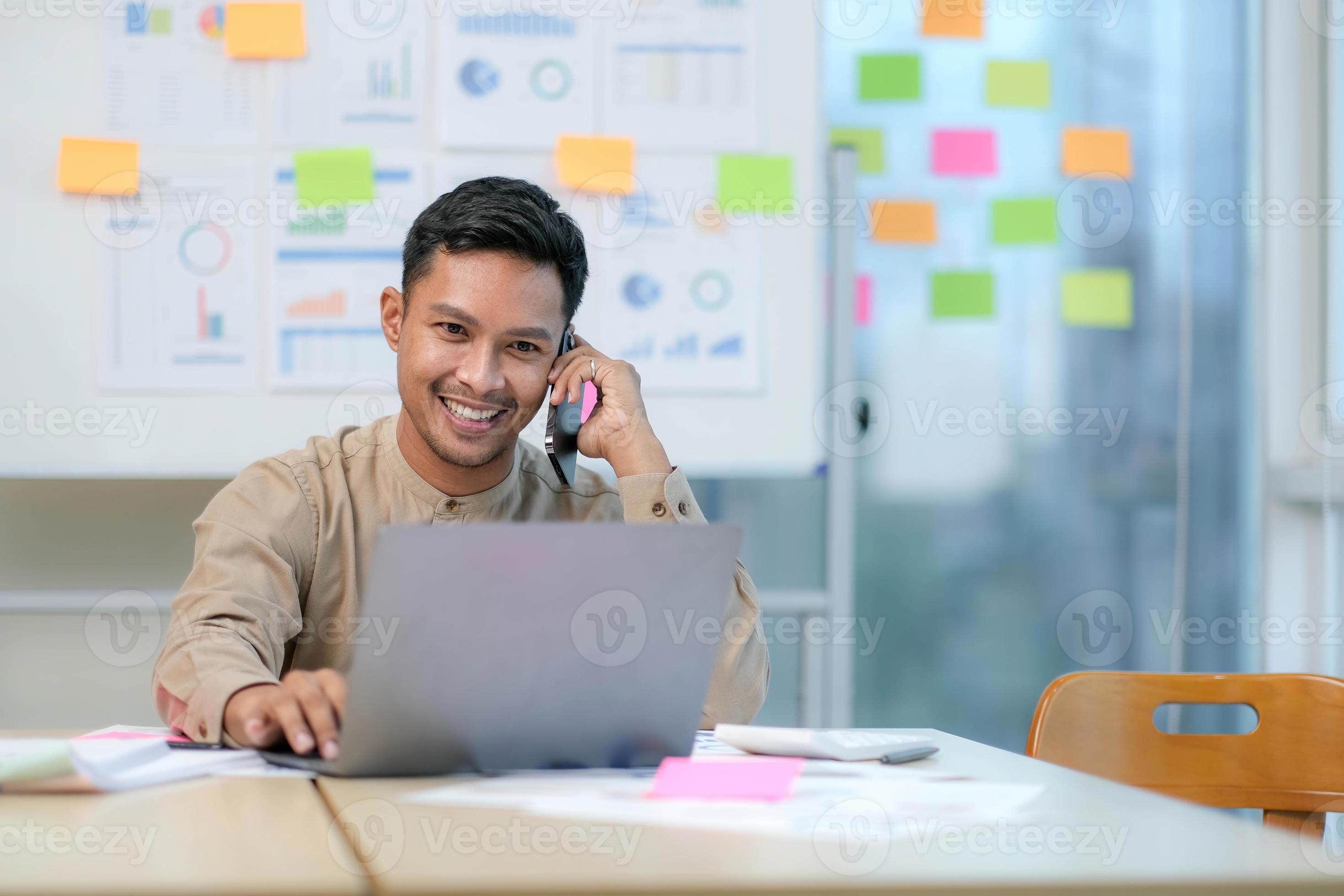 handsome young employee sitting at desk and looking at computer monitor while answering call in ...