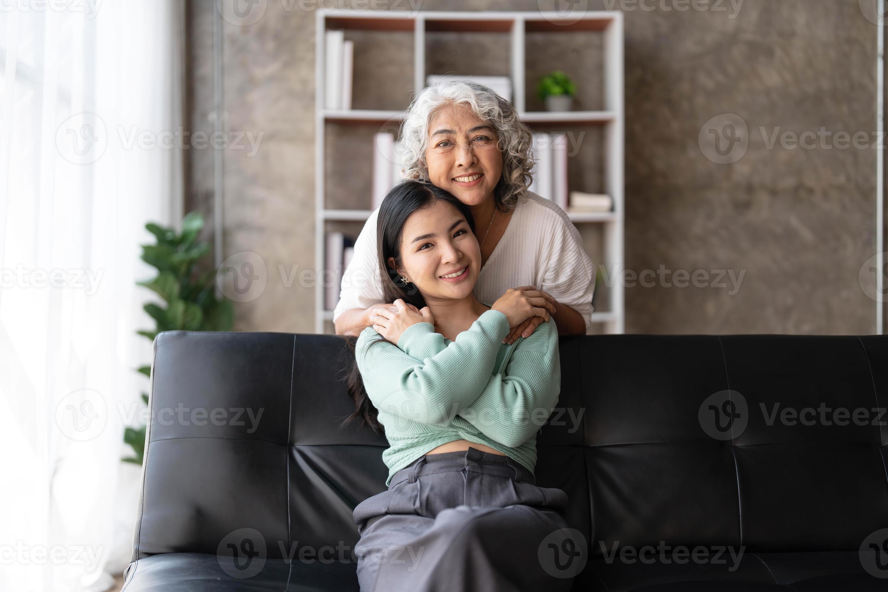 Loving adult daughter hugging older mother, standing behind couch at home, family enjoying ...
