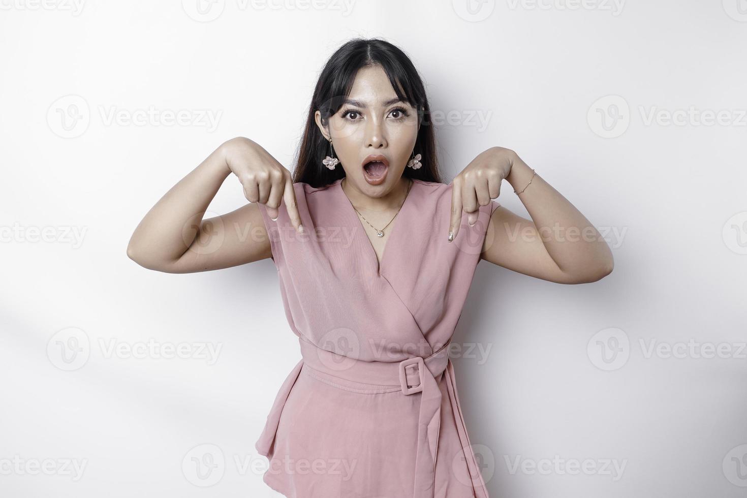 Shocked Asian woman dressed in pink, pointing at the copy space below ...