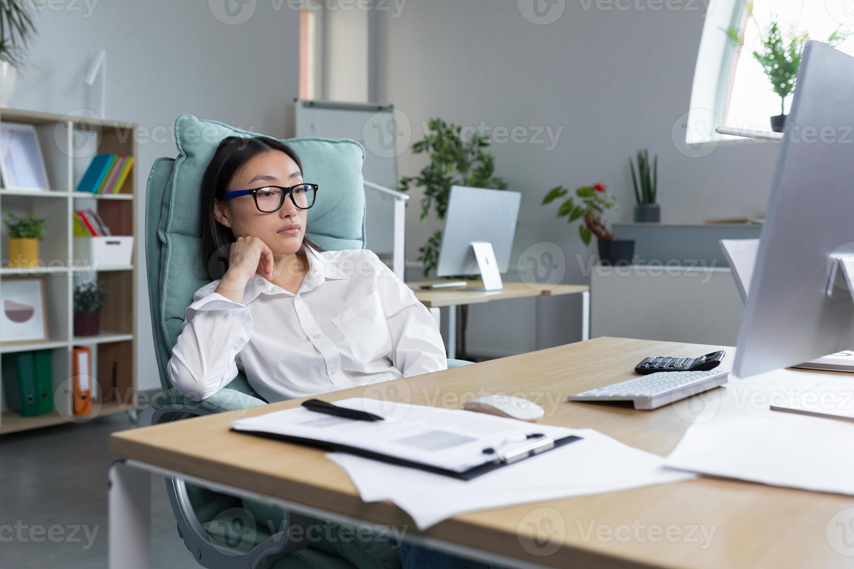 Pensive and sad young Asian businesswoman sitting in the office at the desk in a big chair ...