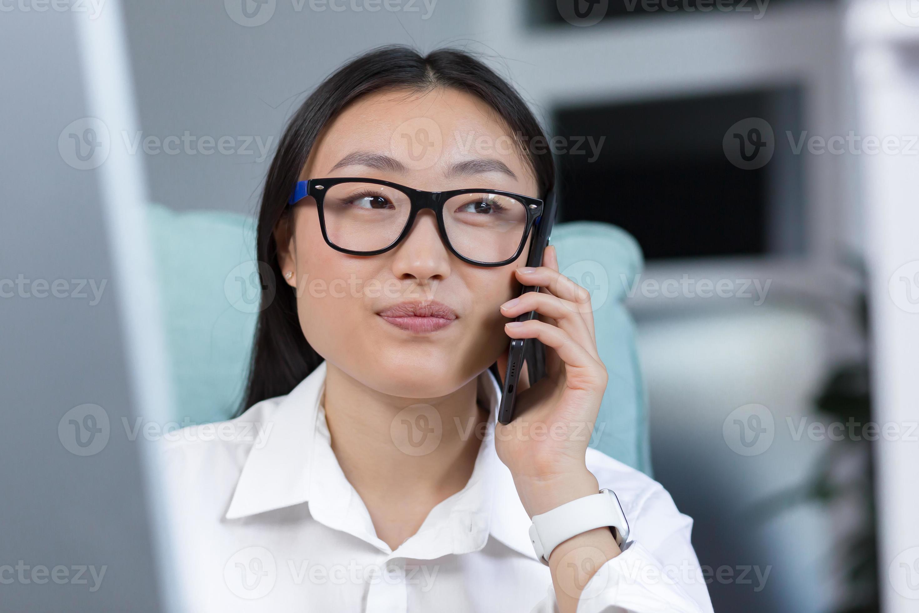 Portrait of a young beautiful Asian business woman in glasses in a white shirt talking phone ...