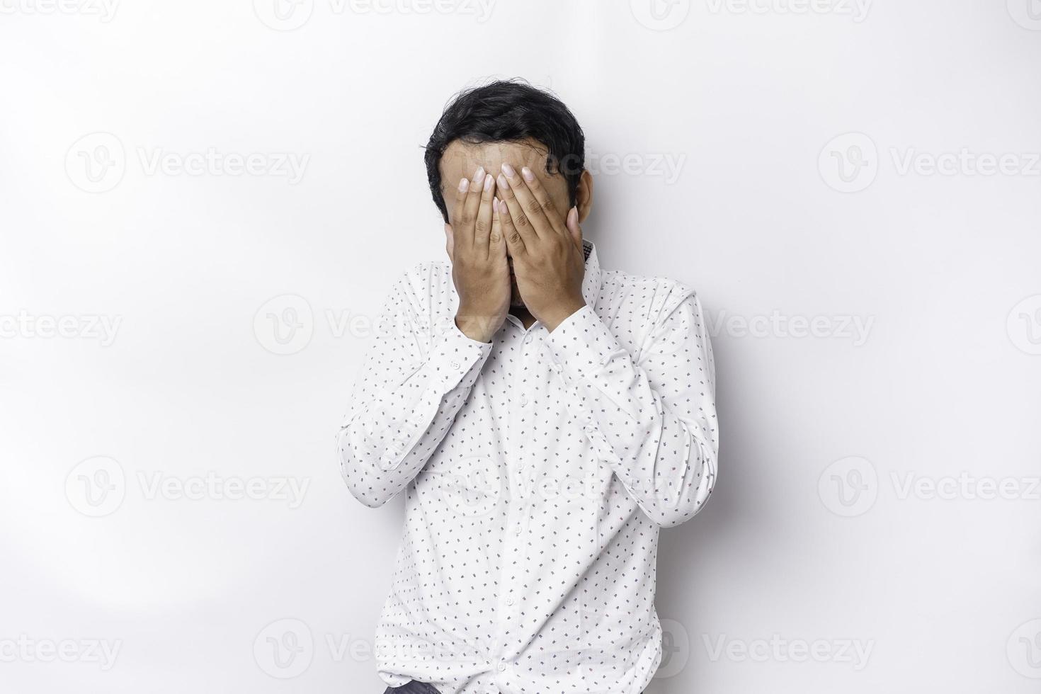 Young scared Asian man isolated on white background, looks depressed, face covered by fingers ...