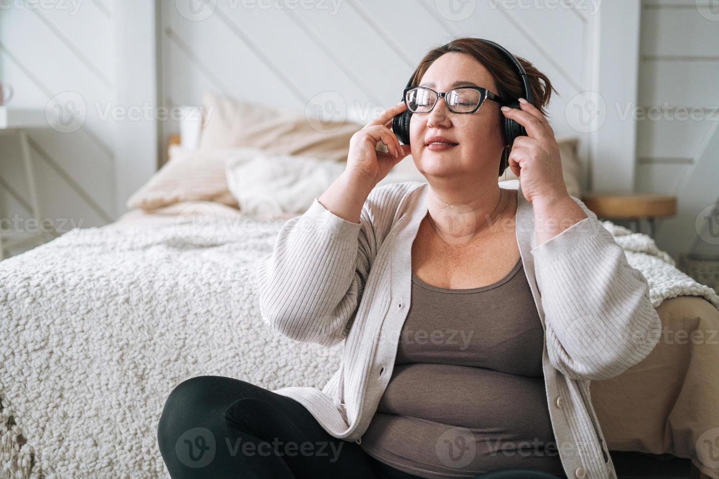 Middle aged plus size smiling brunette woman in home wear listening music in headphones in ...