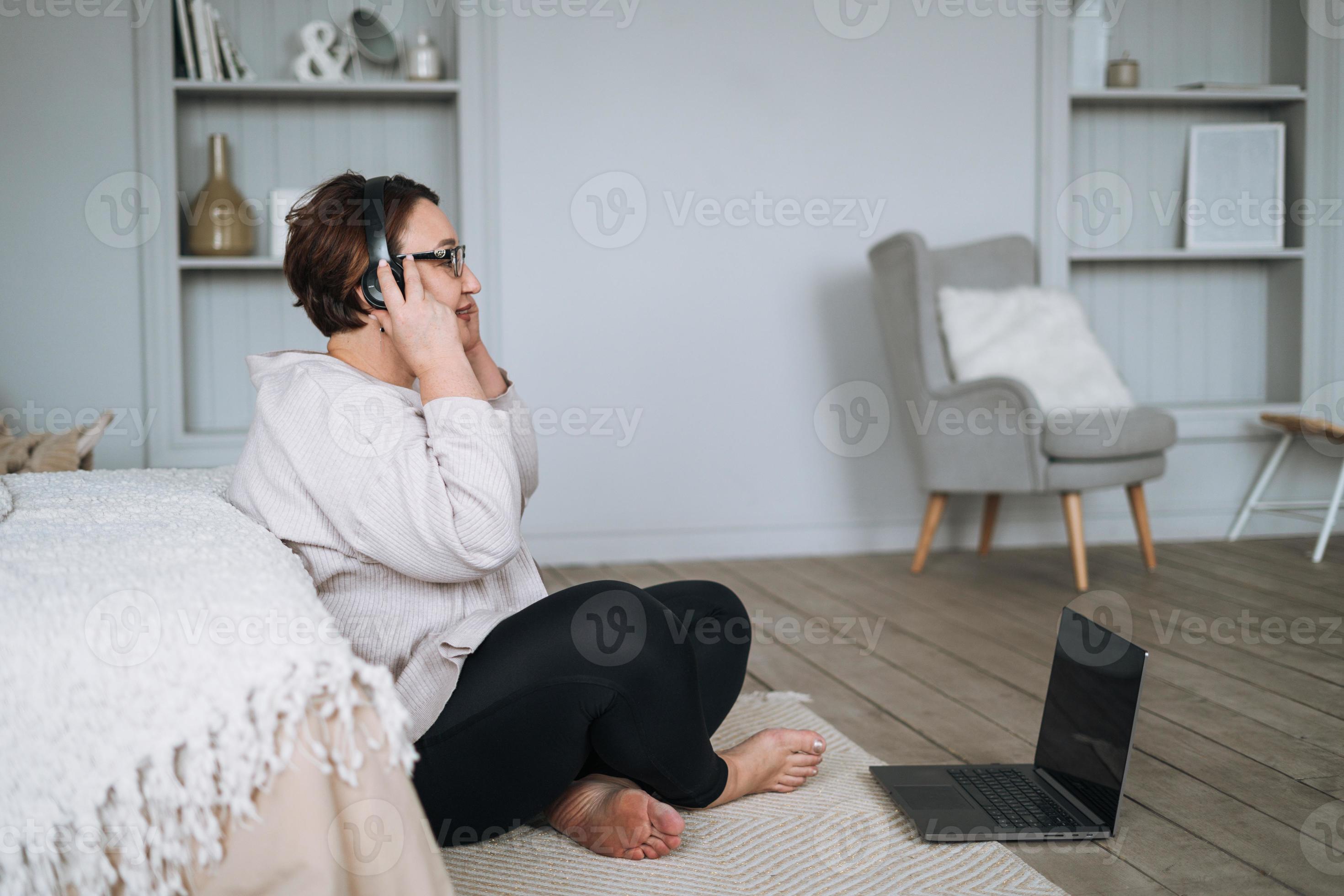Middle aged plus size smiling brunette woman in home wear listening music in headphones using ...