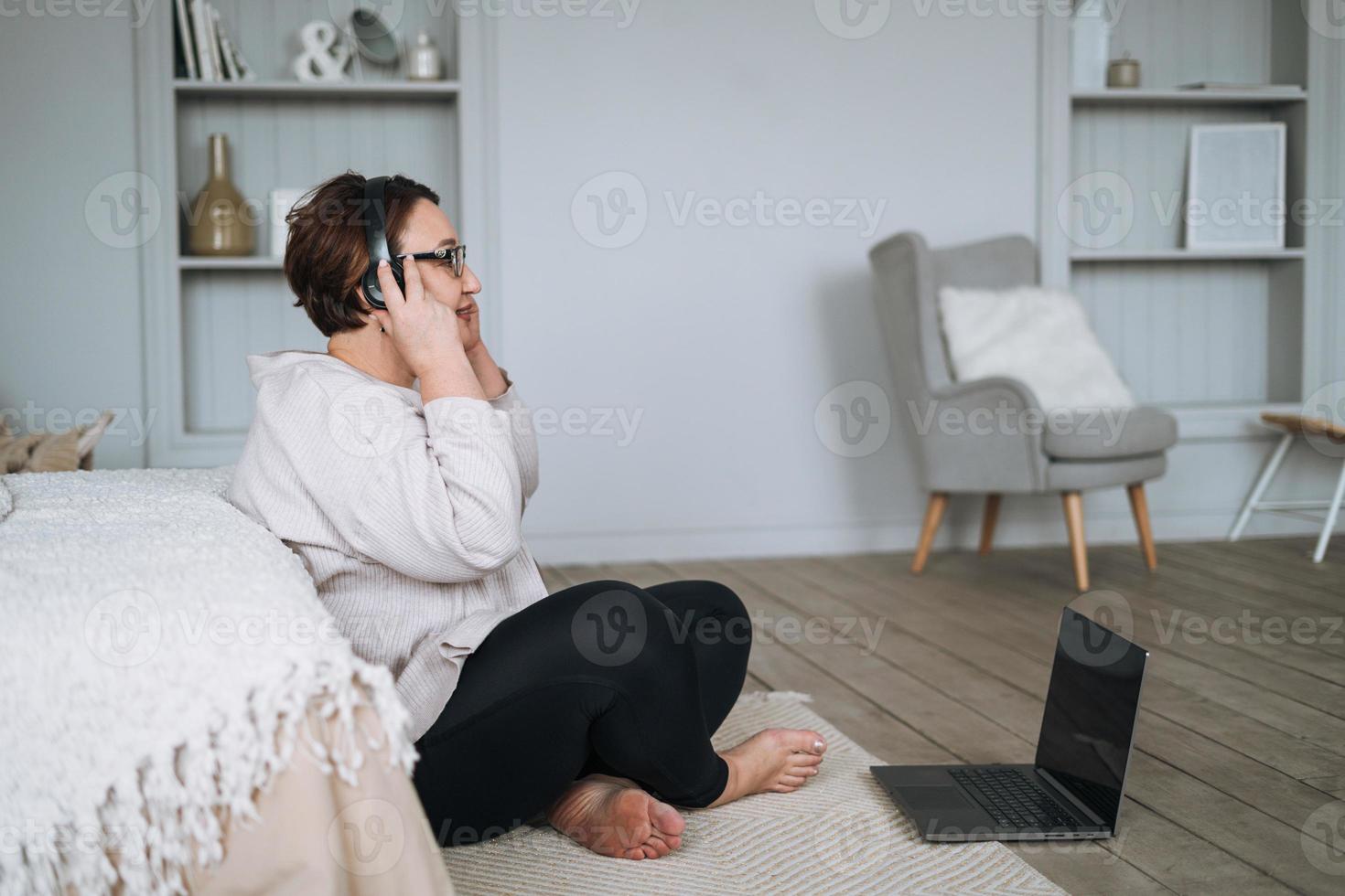 Middle aged plus size smiling brunette woman in home wear listening music in headphones using ...