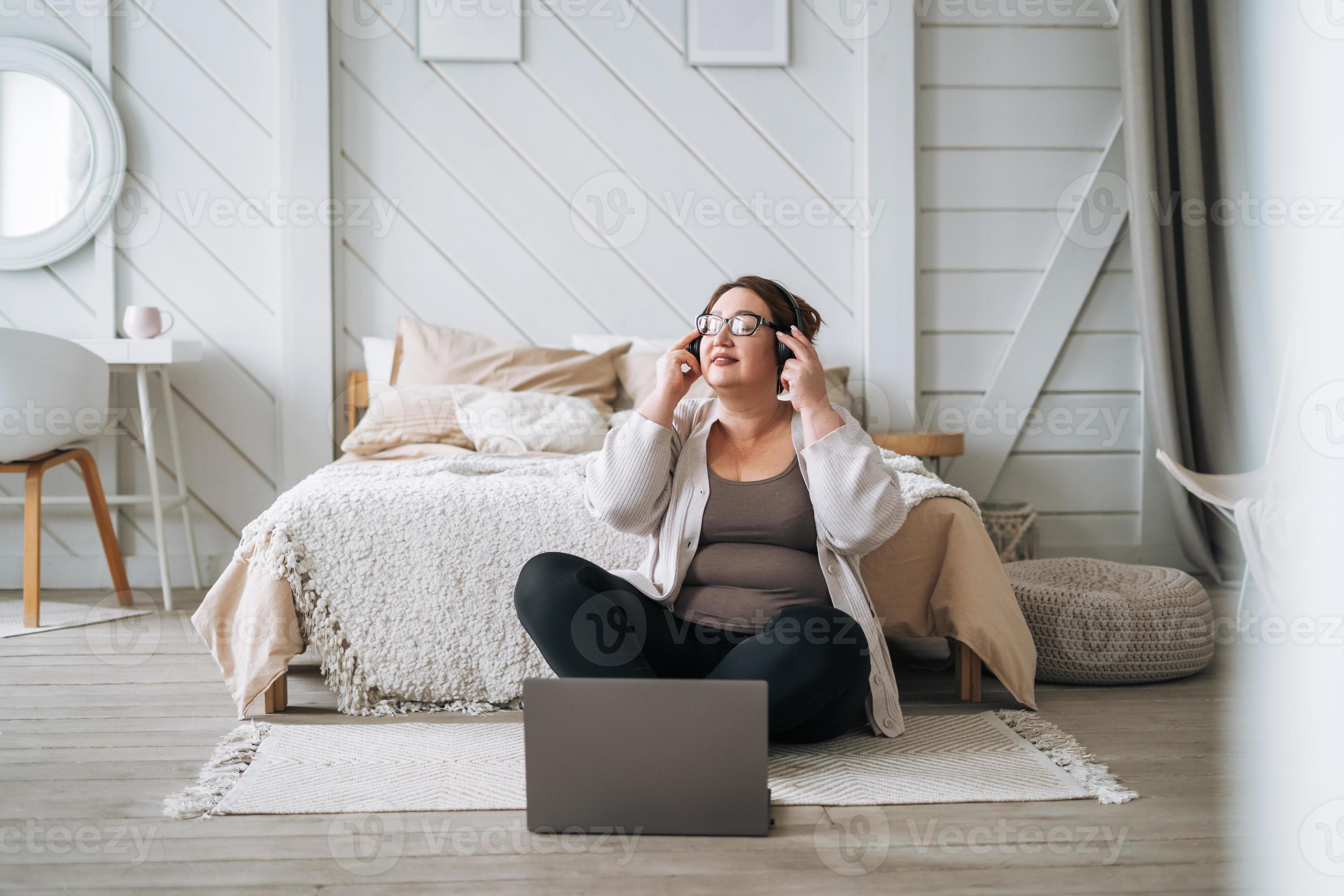 Middle aged plus size smiling brunette woman in home wear listening music in headphones using ...