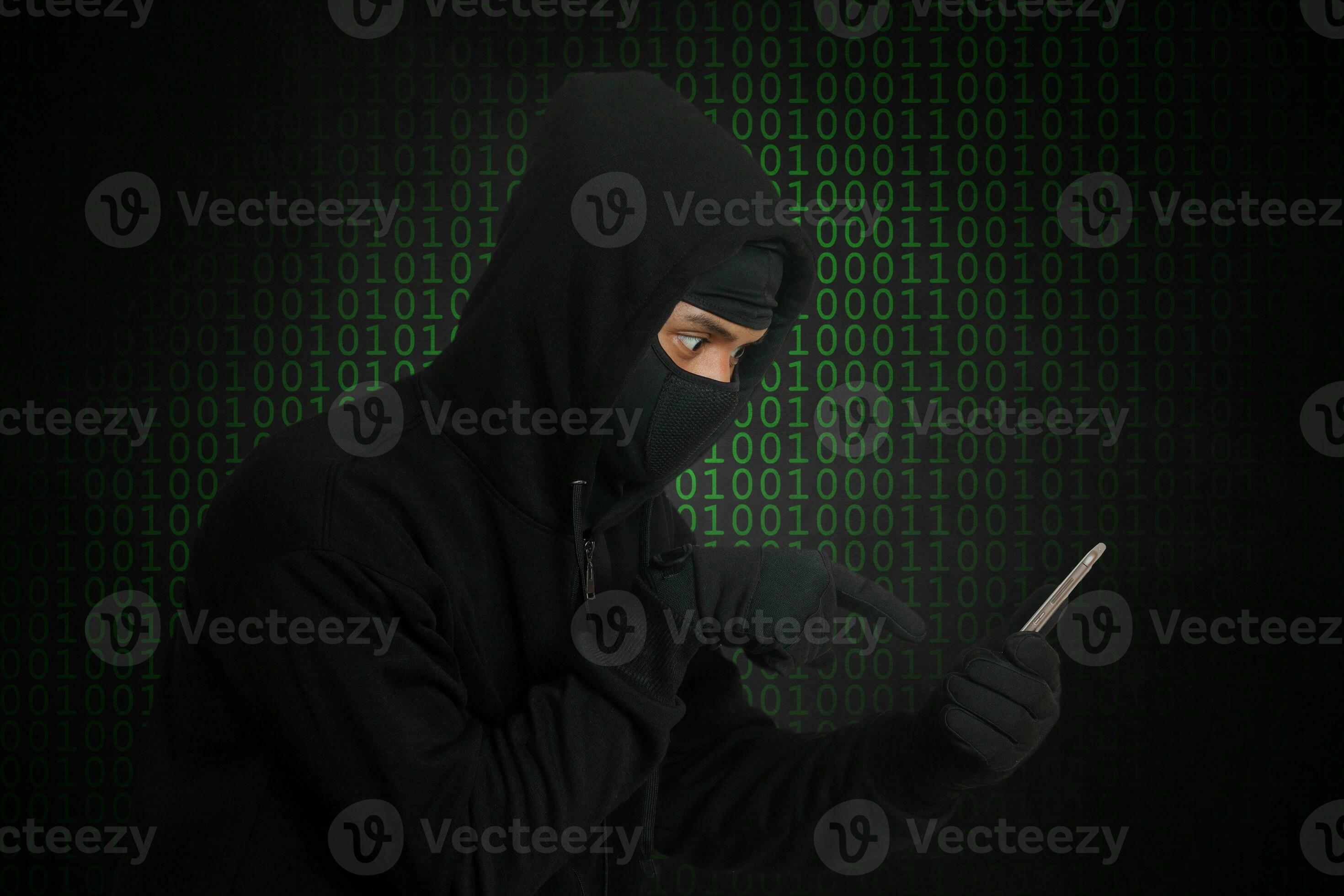 Portrait Of Mysterious Man Wearing Black Hoodie And Mask Doing Hacking Activity On Mobile Phone