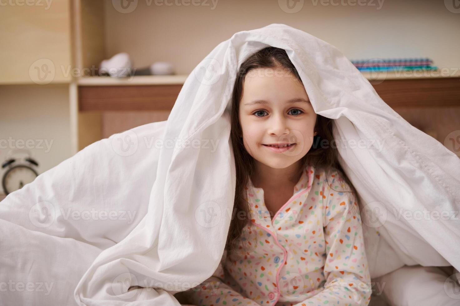 Caucasian happy little child girl sitting on comfortable bed and peeking out from under soft ...