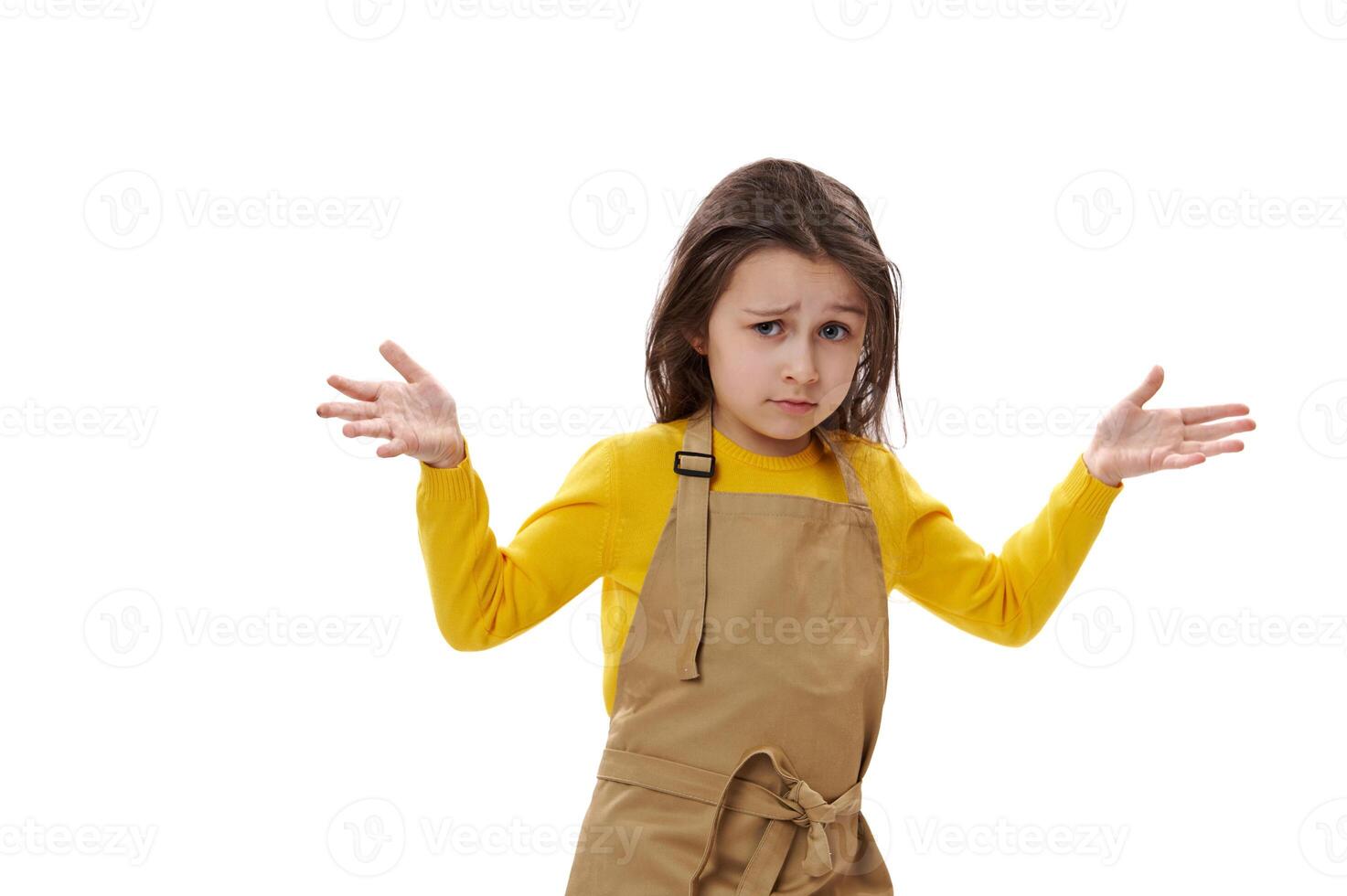 Amazed little child girl in beige chef's apron, asking questions, holding hands palms up ...